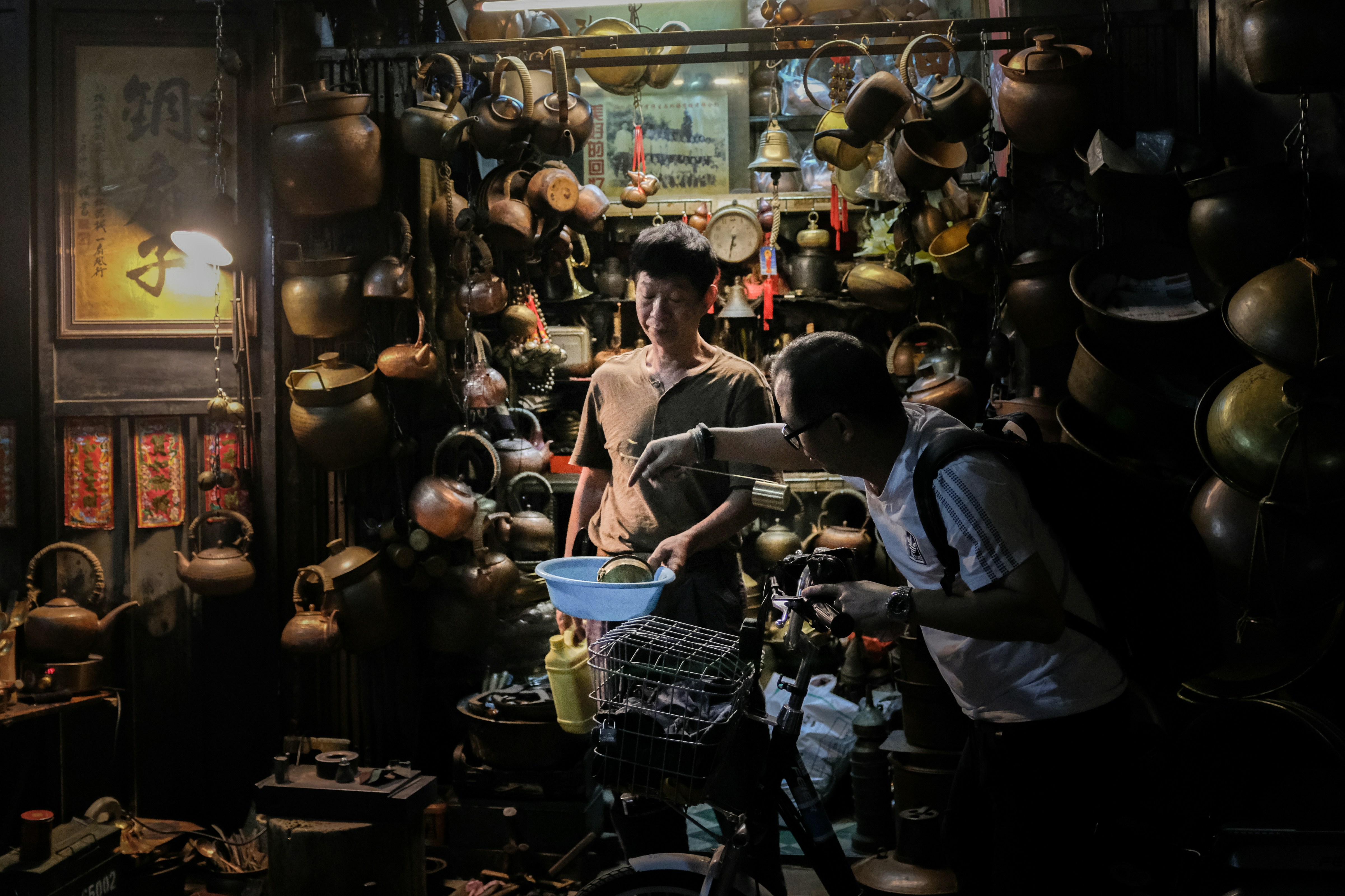 Dimly lit shop filled with hanging metal pots and artifacts, where two men engage in conversation.