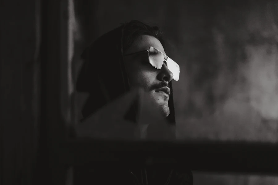 Black-and-white portrait of a man in sunglasses and a hood, lit softly from the side with a blurred background. Framed through dark foreground shapes for a moody, cinematic feel.