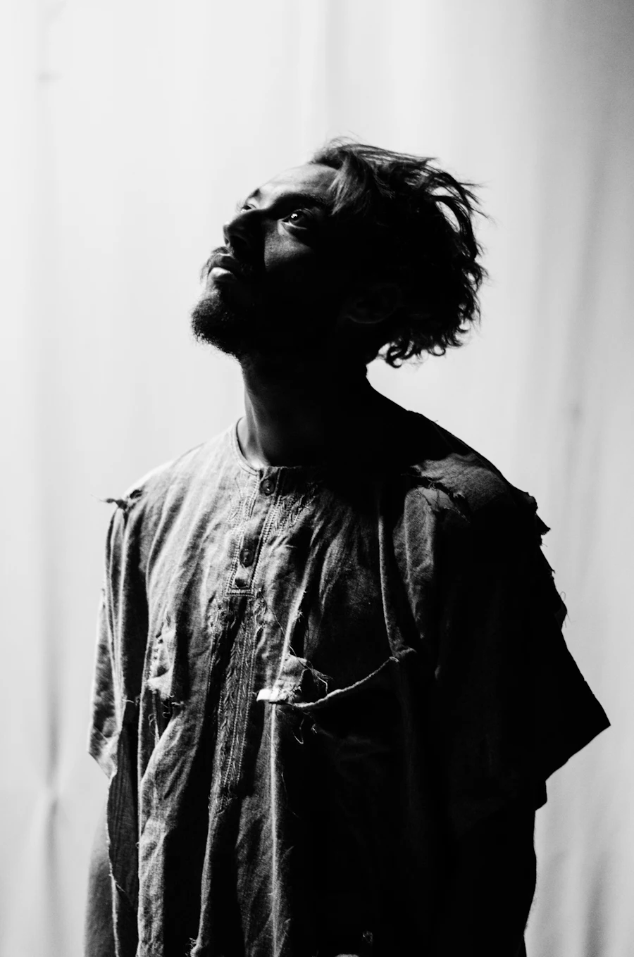 High-contrast black-and-white portrait of a man looking upward, his face mostly in shadow against a bright background. His textured shirt and tousled hair add a dramatic, moody feel.