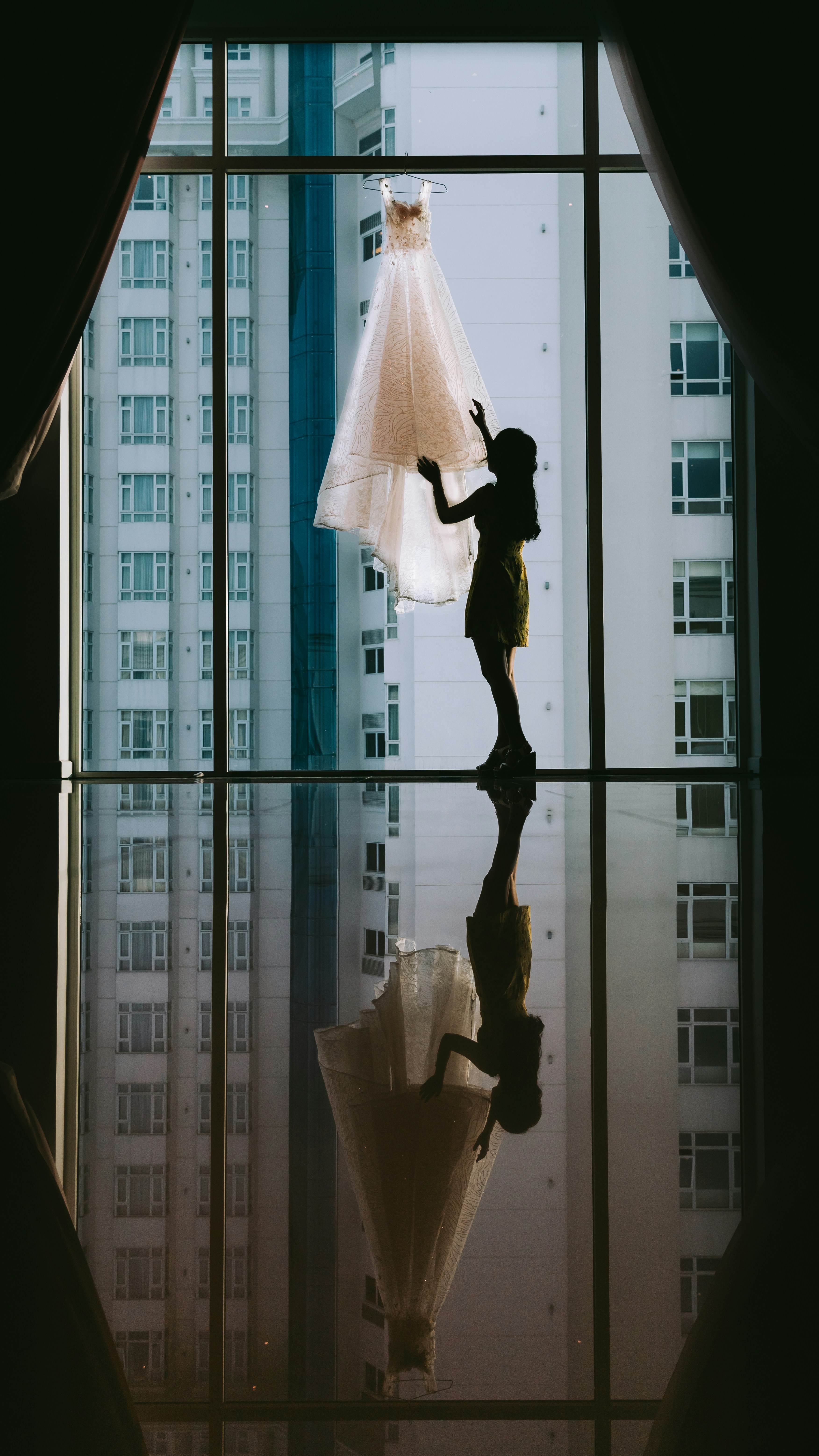 A silhouetted figure holds a wedding dress against a backdrop of modern architecture, with the reflection creating a dreamlike effect.