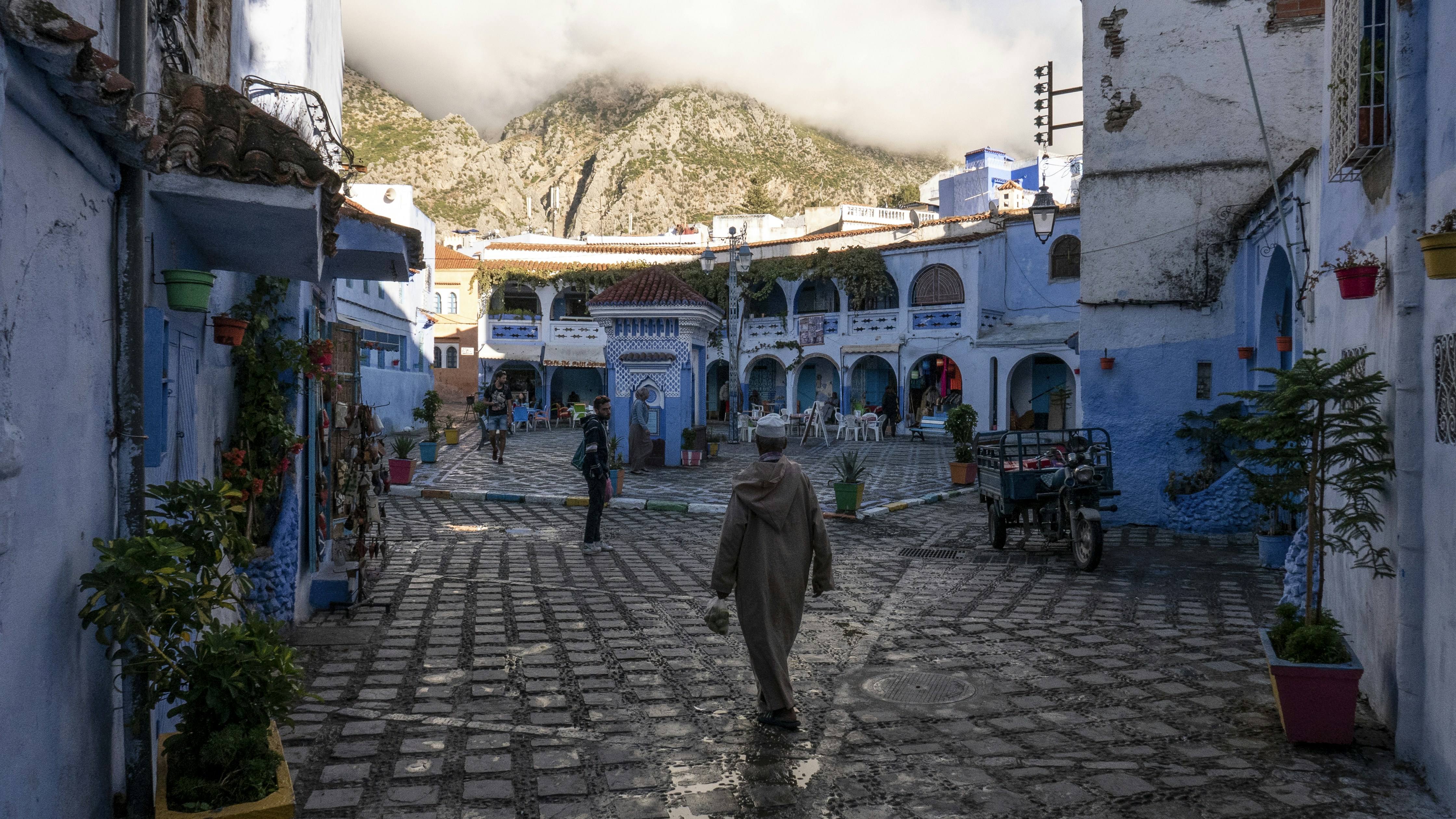 A small cozy square hidden in the hearth of Chefchaouen. A stunning view on cloudy mountains on the background and in the foreground a man wearing traditional moroccan dresses is walking into the square. | people walking on street in between concrete buildings during daytime