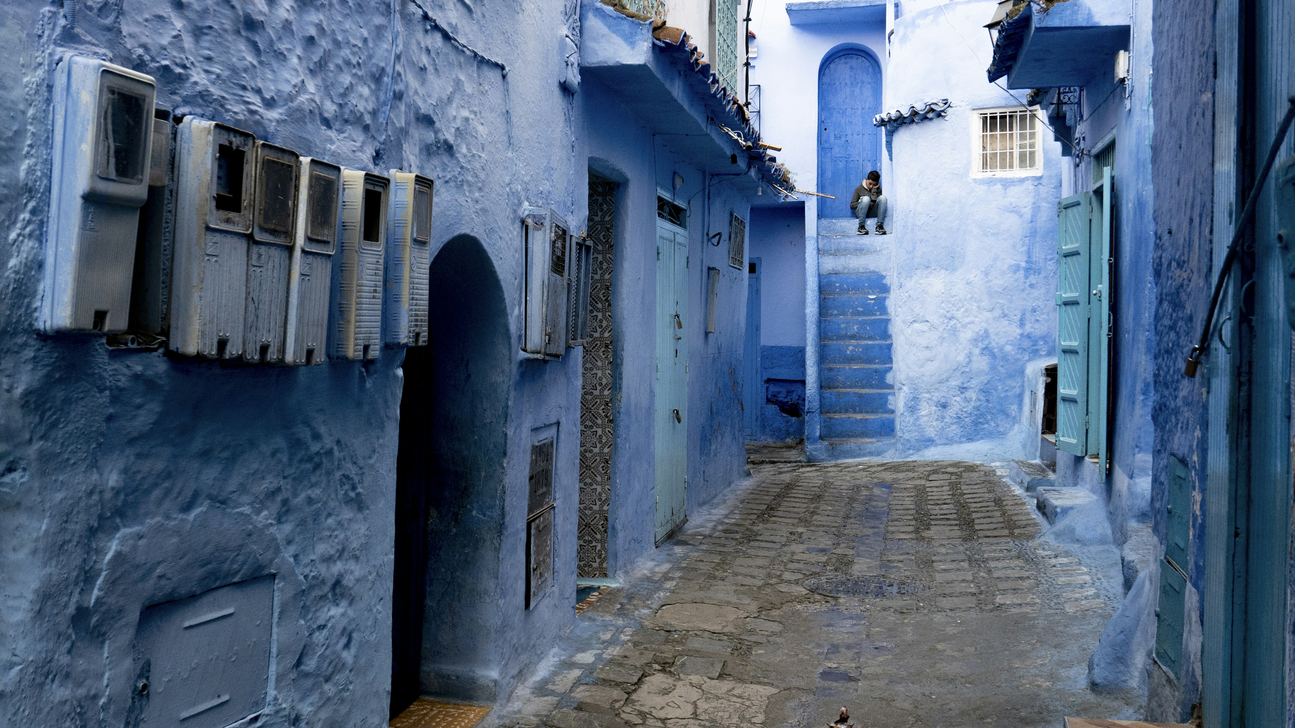 Narrow alleyway adorned in varying shades of blue, featuring a lone figure seated on stairs. The scene captures the tranquility of a quiet moment in a vibrant setting.