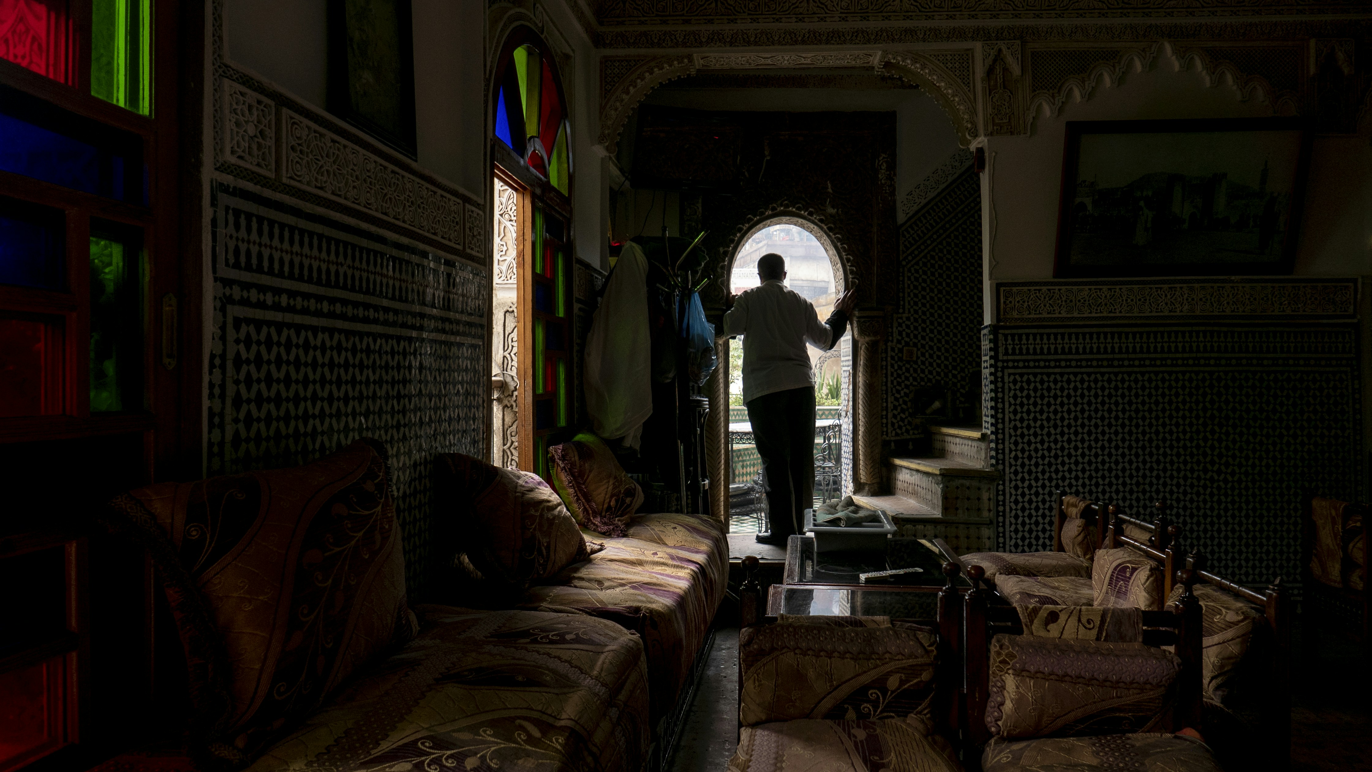 Silhouette of a person standing by a brightly lit balcony door in a dimly lit Moroccan bar.