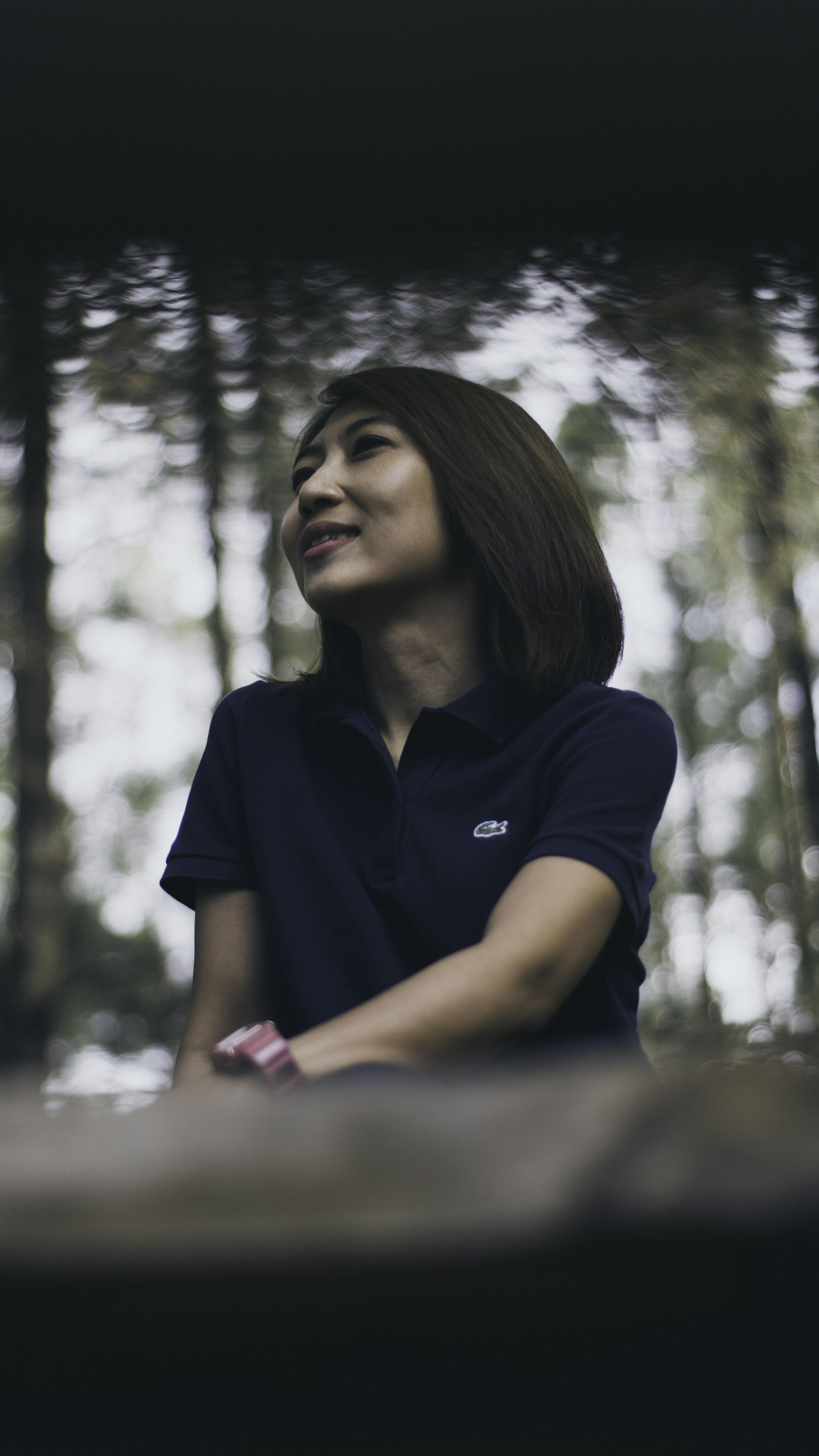 Portrait photograph of a woman in a navy polo shirt sitting outdoors among trees, with shallow depth of field producing a soft, blurred background.