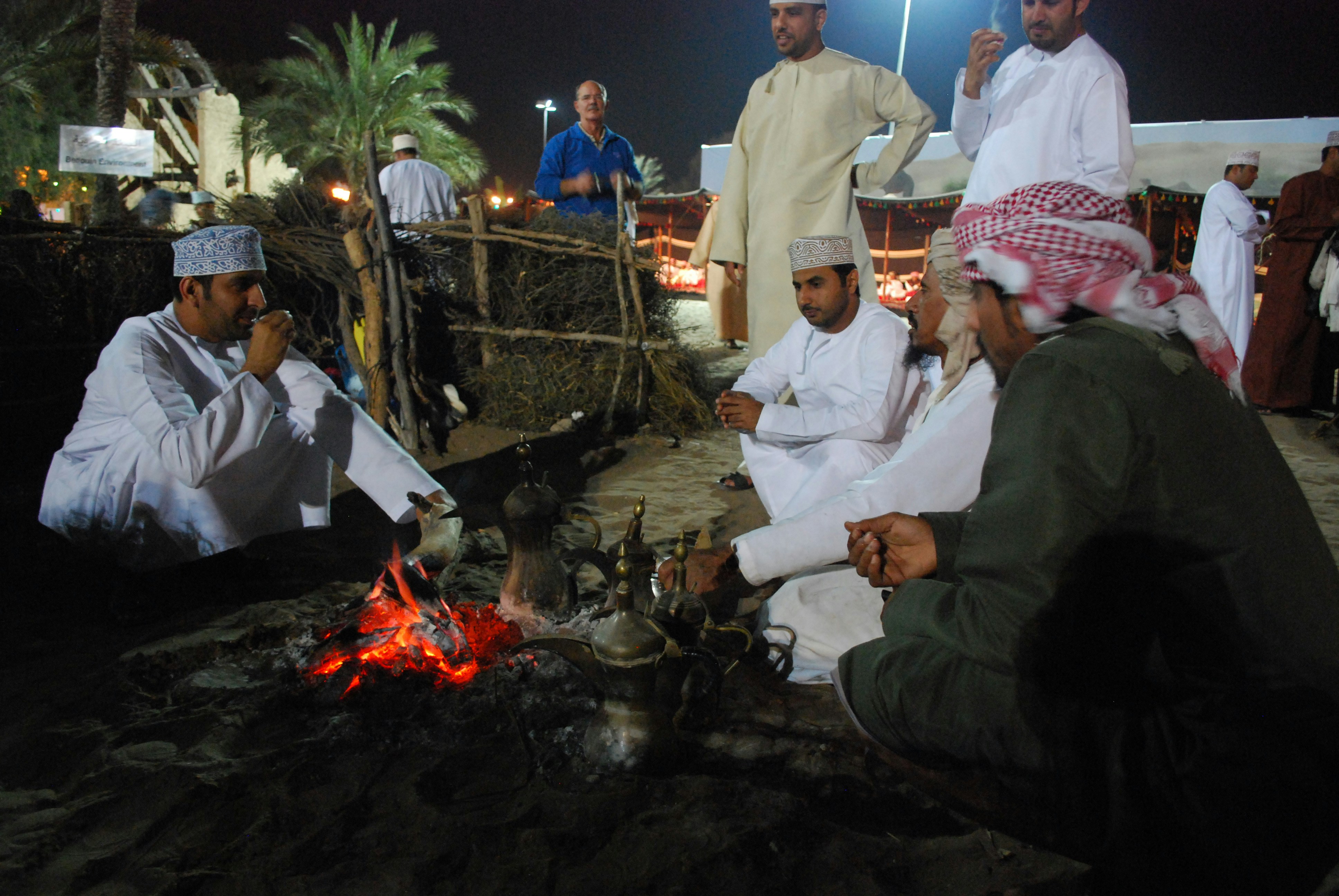 People in traditional attire seated around a bonfire with coffee pots, under a starlit sky.
