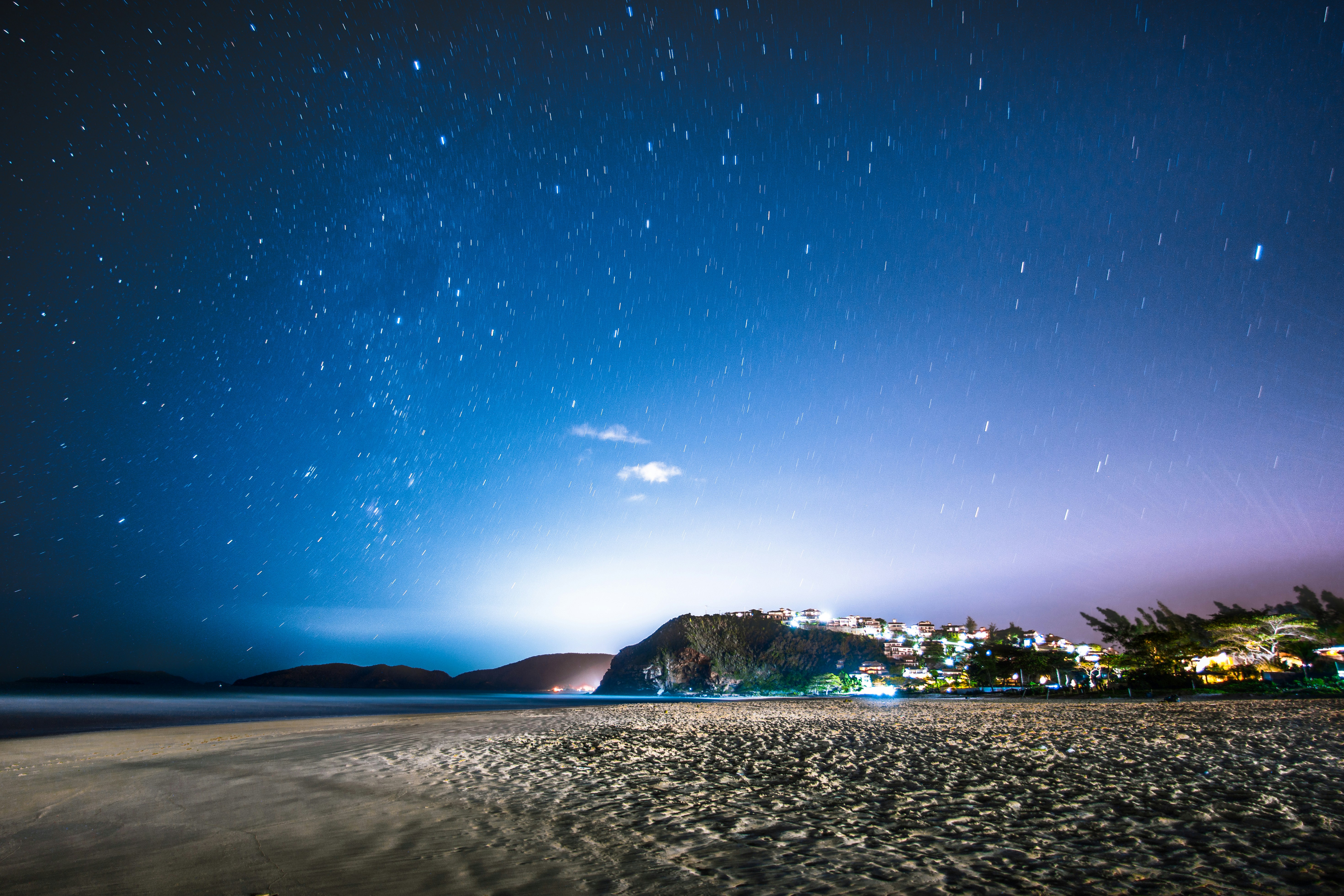 Long exposure of a starry sky over a tranquil beach at night with distant city lights.