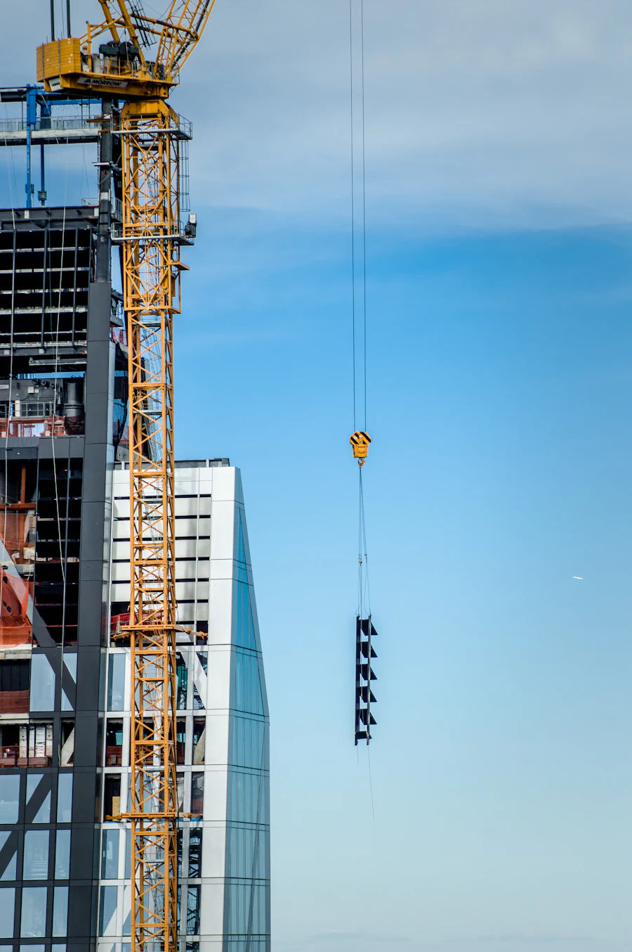 Construction crane and building under a bright blue sky; the suspended load and crane cables are visible against the façade, illustrating urban development and scale.