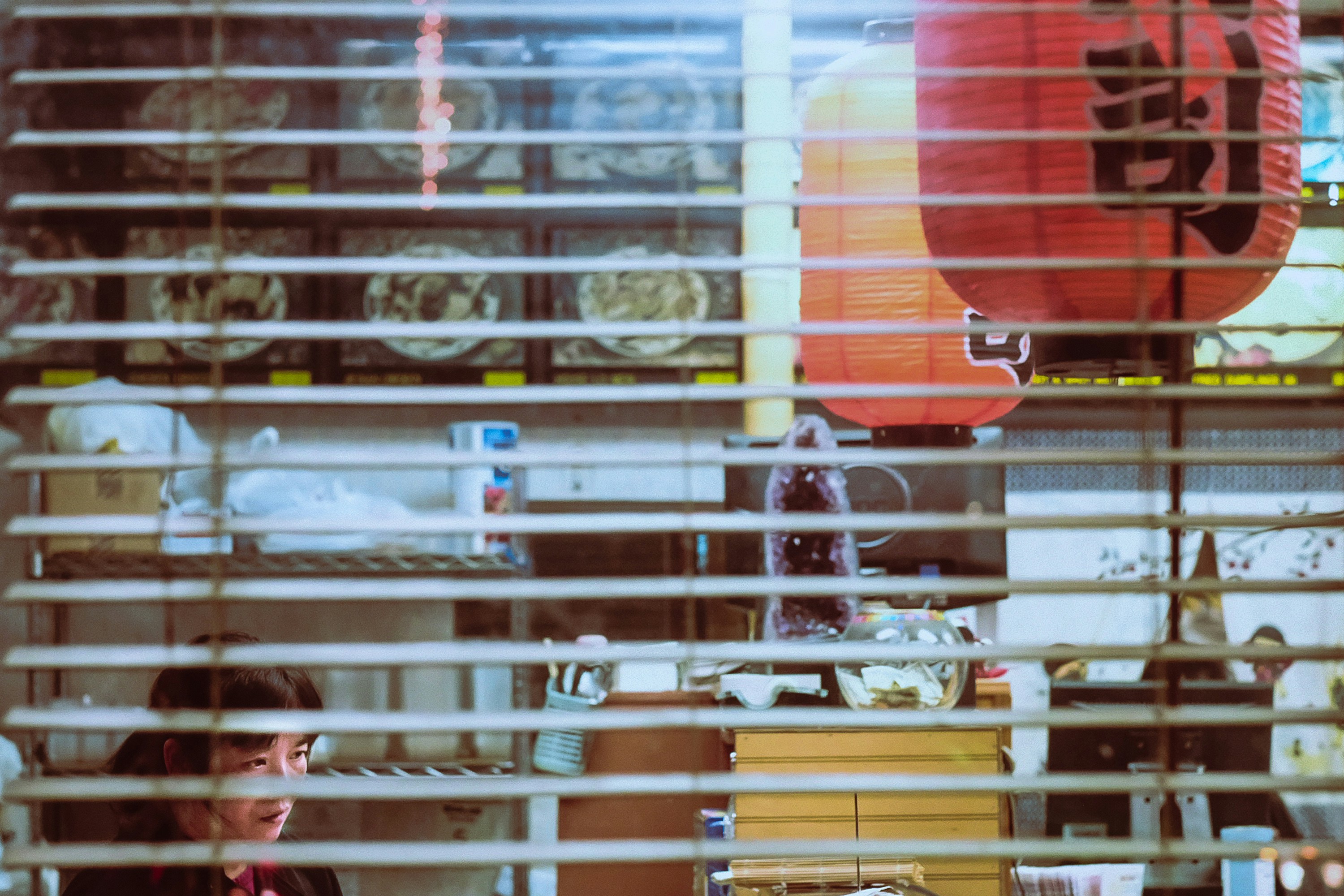 Person seated inside a restaurant, partially obscured by horizontal blinds and illuminated by red lanterns.