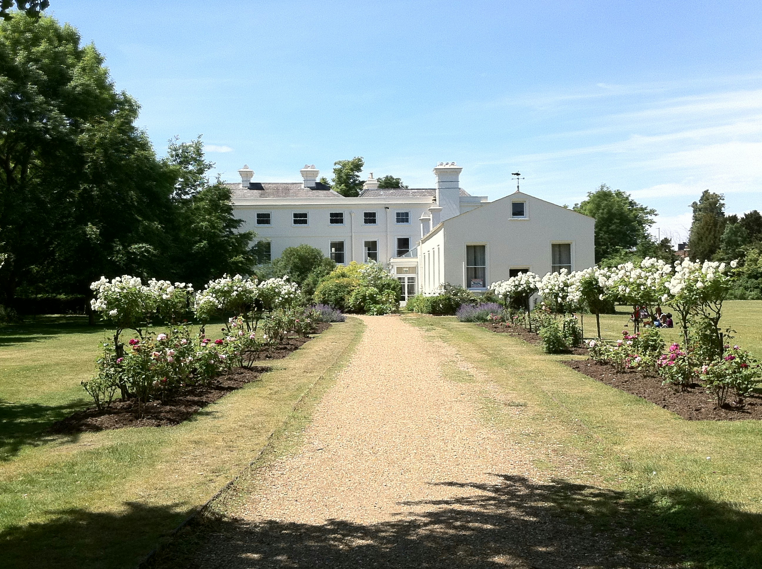 Sunlit gravel path leads through a rose-bordered garden to a white estate, with neatly trimmed lawns and flowering bushes on both sides.