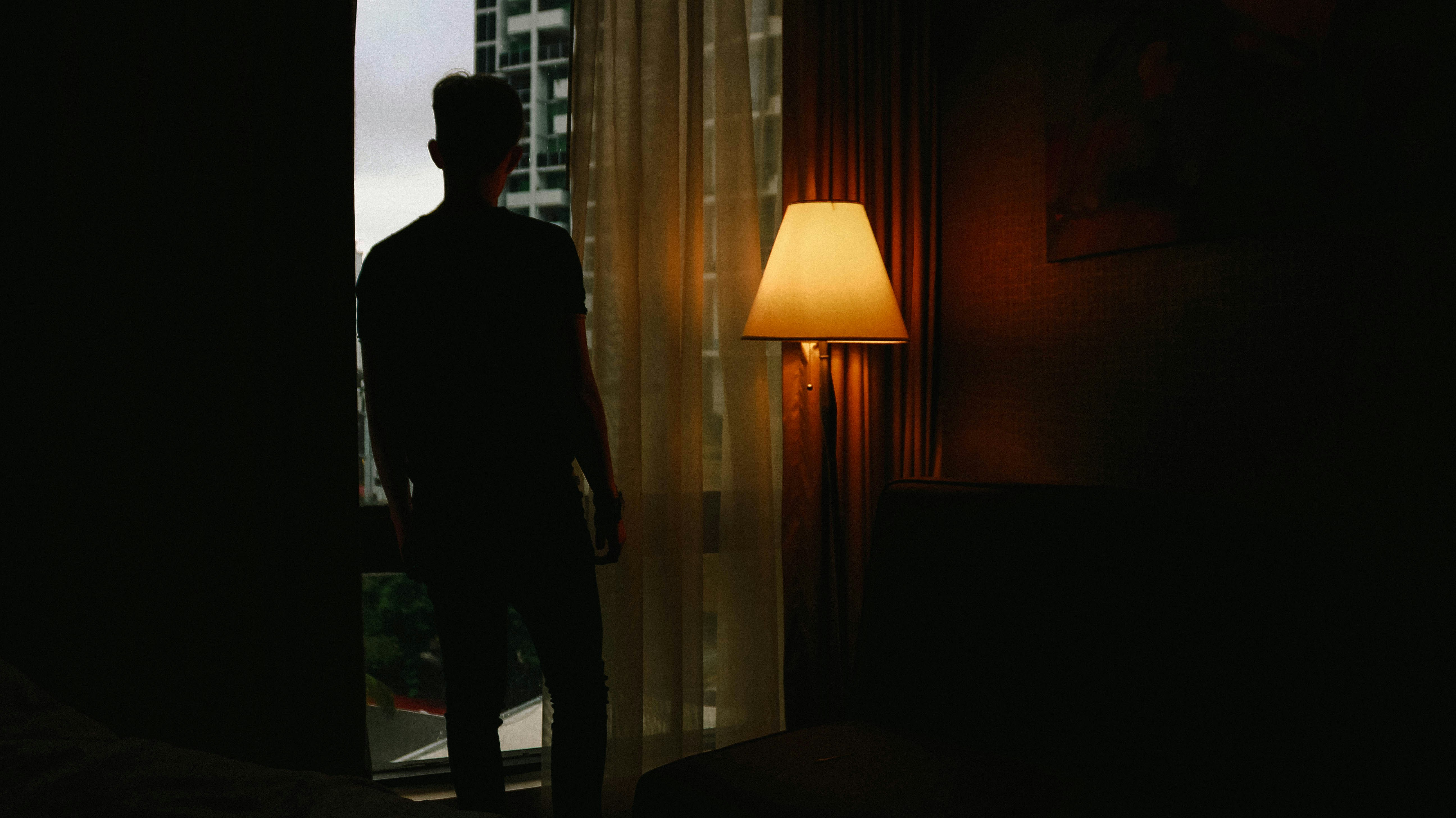 A silhouette stands by a window, gazing out at the cityscape beyond sheer curtains, illuminated by the warm glow of a nearby lamp.
