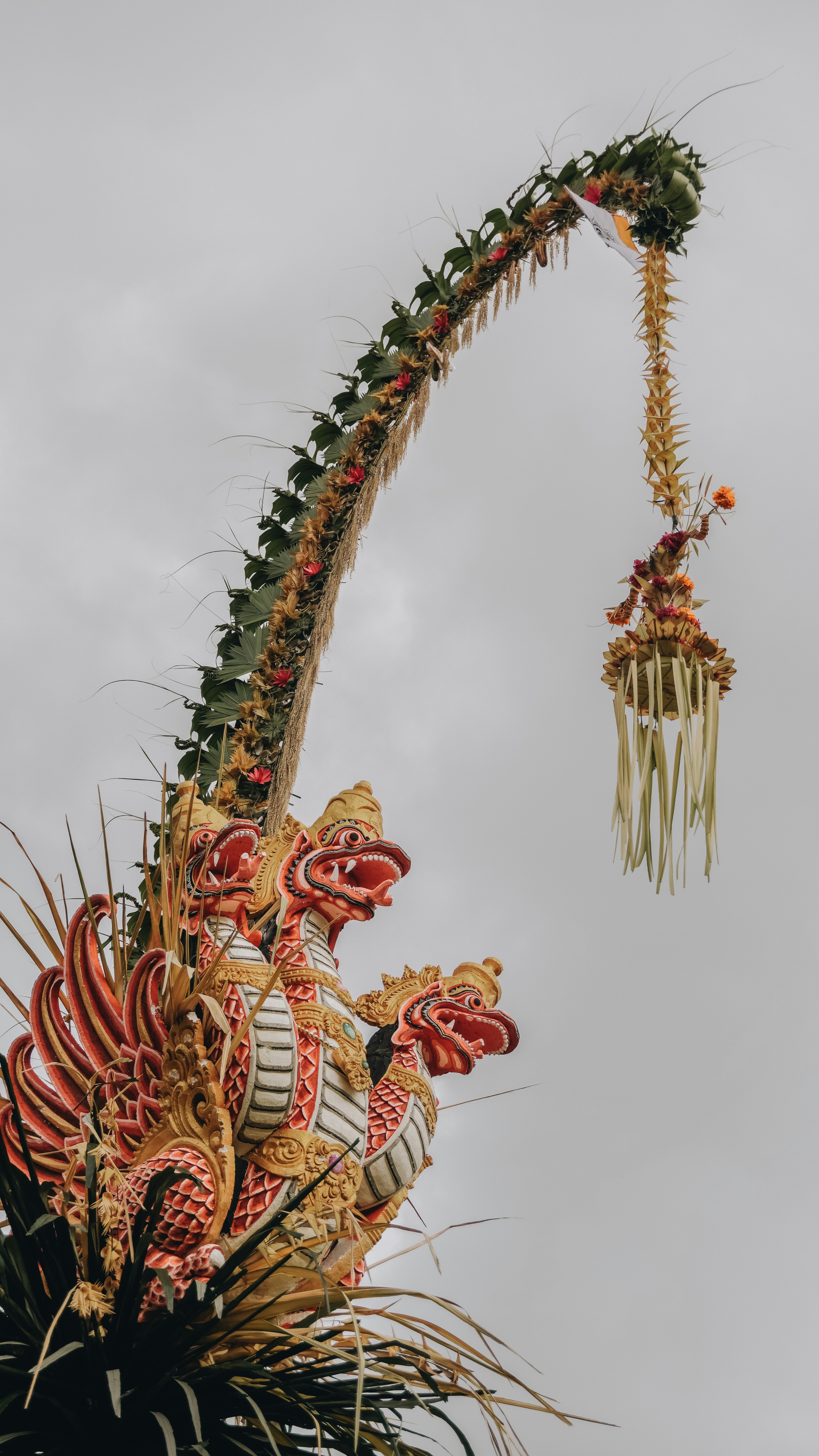 Traditional Balinese penjor adorned with intricate carvings and floral decorations, set against a grey sky.