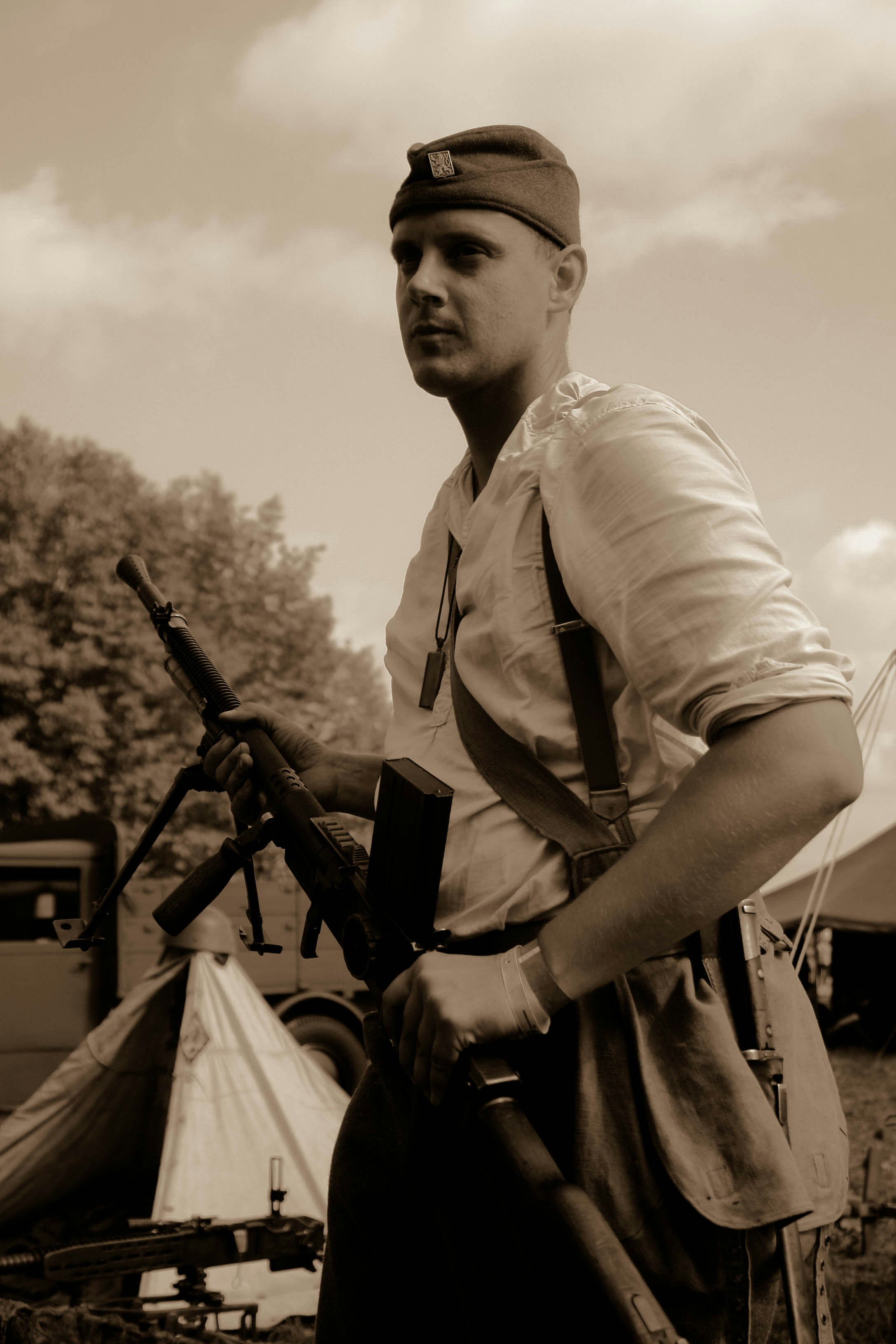 Vintage-toned portrait of a soldier in period attire, holding a rifle with a camp backdrop including tents and a truck.