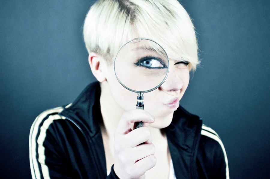 Blonde person winking while holding a magnifying glass over one eye, against a teal background. Black jacket with white stripes frames the portrait.