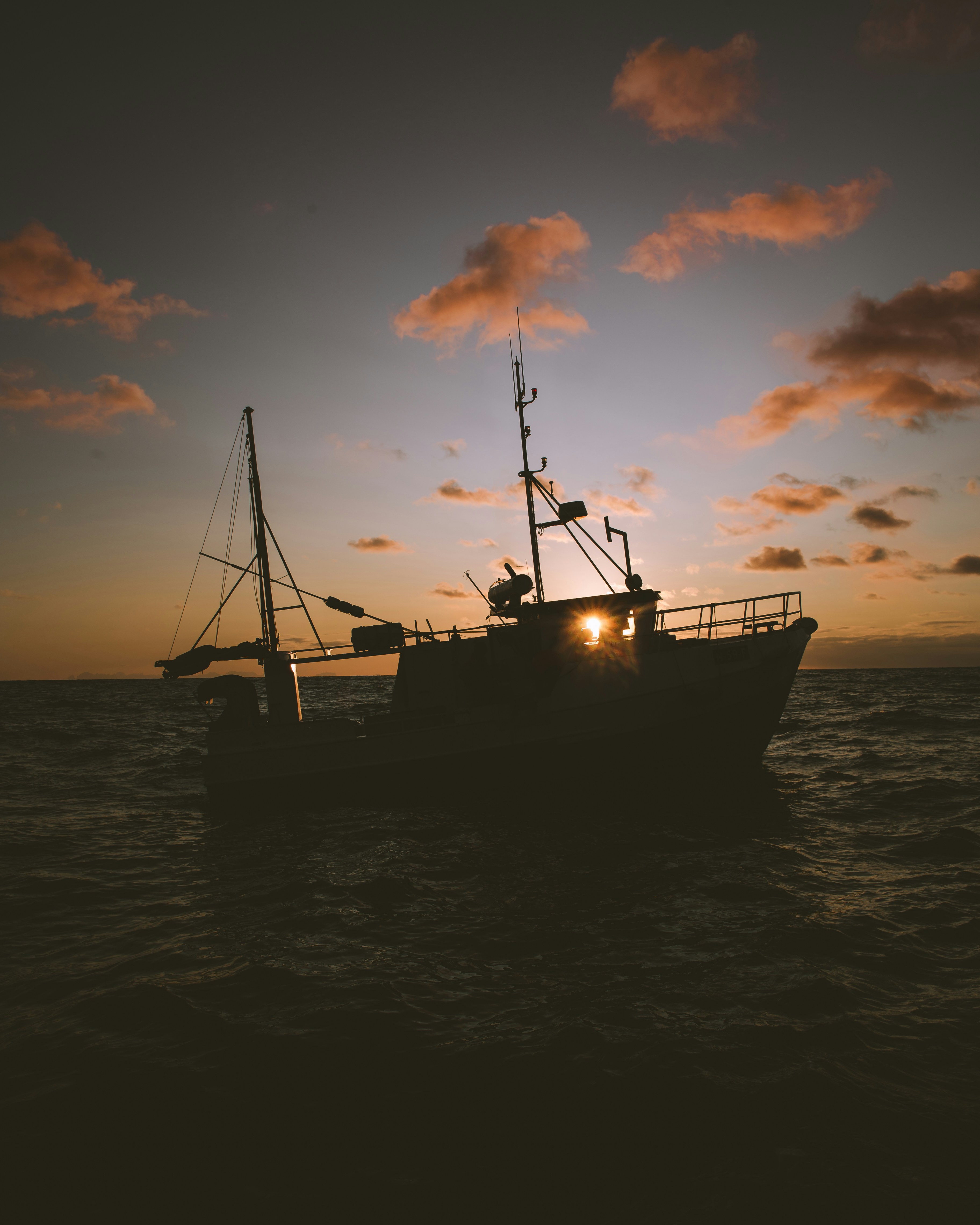 Fishing boat silhouetted against a vibrant sunset sky, with clouds reflecting warm hues over the water's surface.