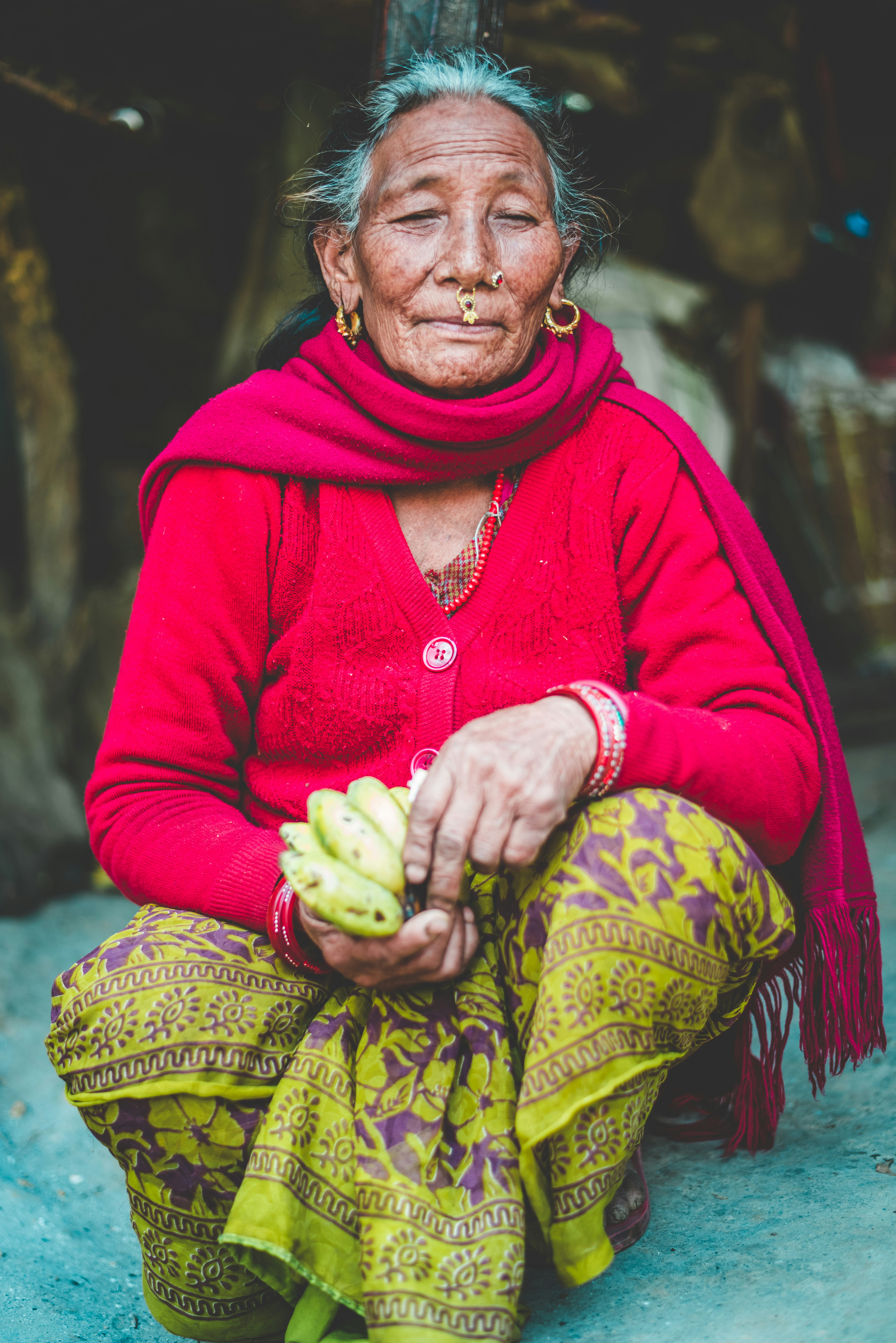 Elderly woman dressed in vibrant red and patterned attire, holding a bunch of bananas, exuding warmth and resilience.