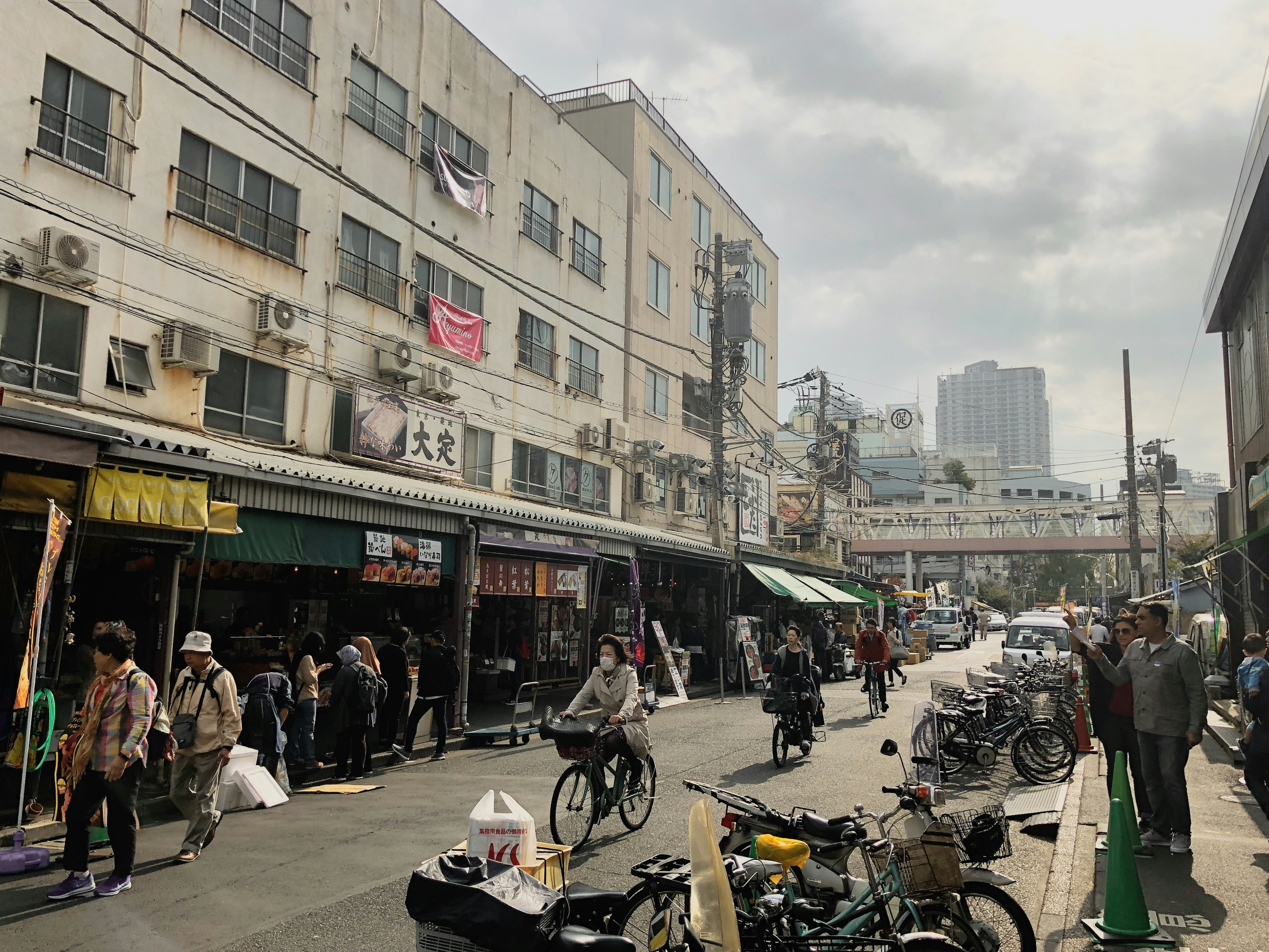 Bustling street scene with pedestrians, cyclists, and shops in Tsukiji Market under a cloudy sky.