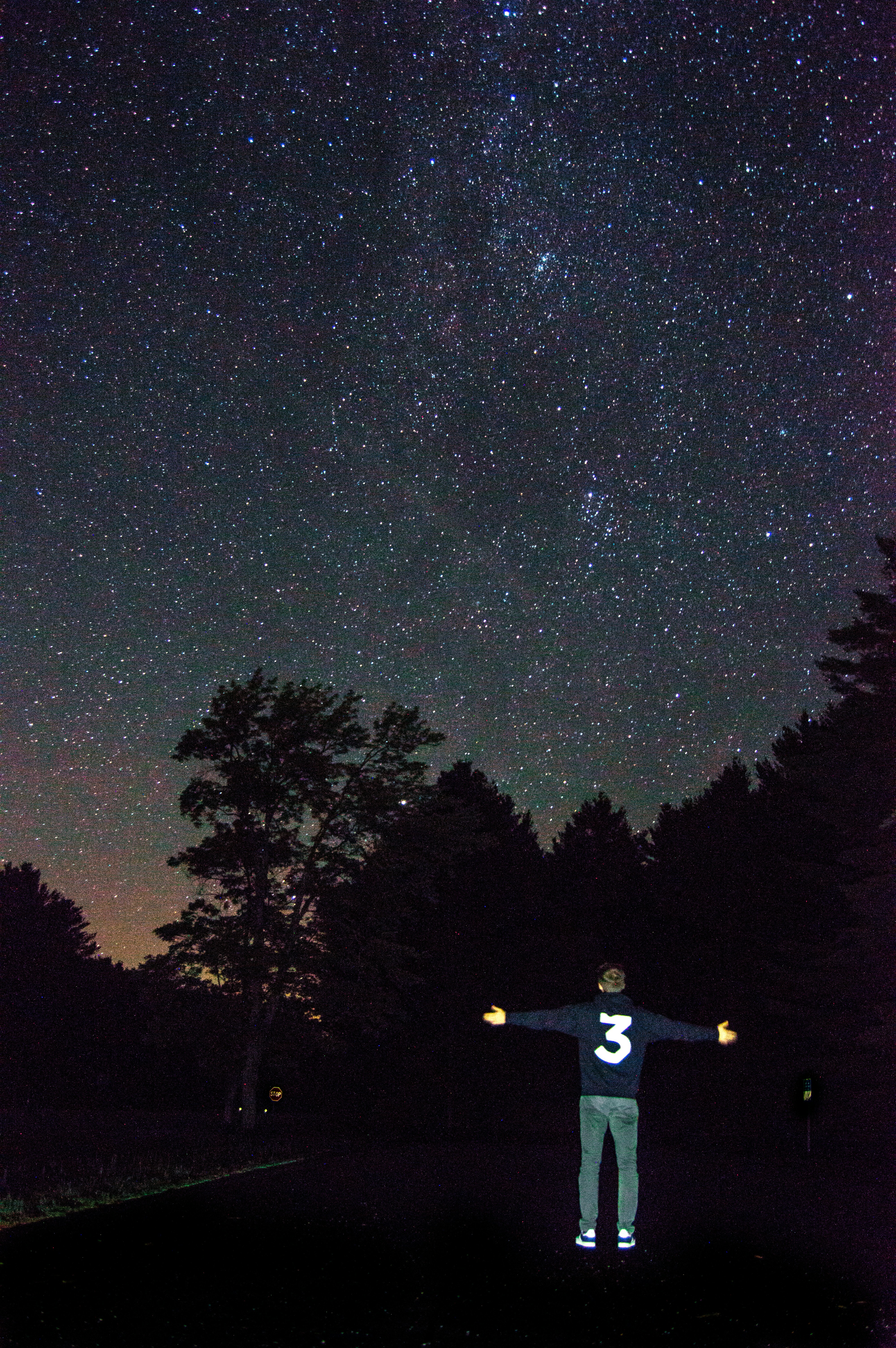 A figure stands with arms outstretched beneath a starry sky, surrounded by silhouettes of trees. The vastness of the universe is on full display.