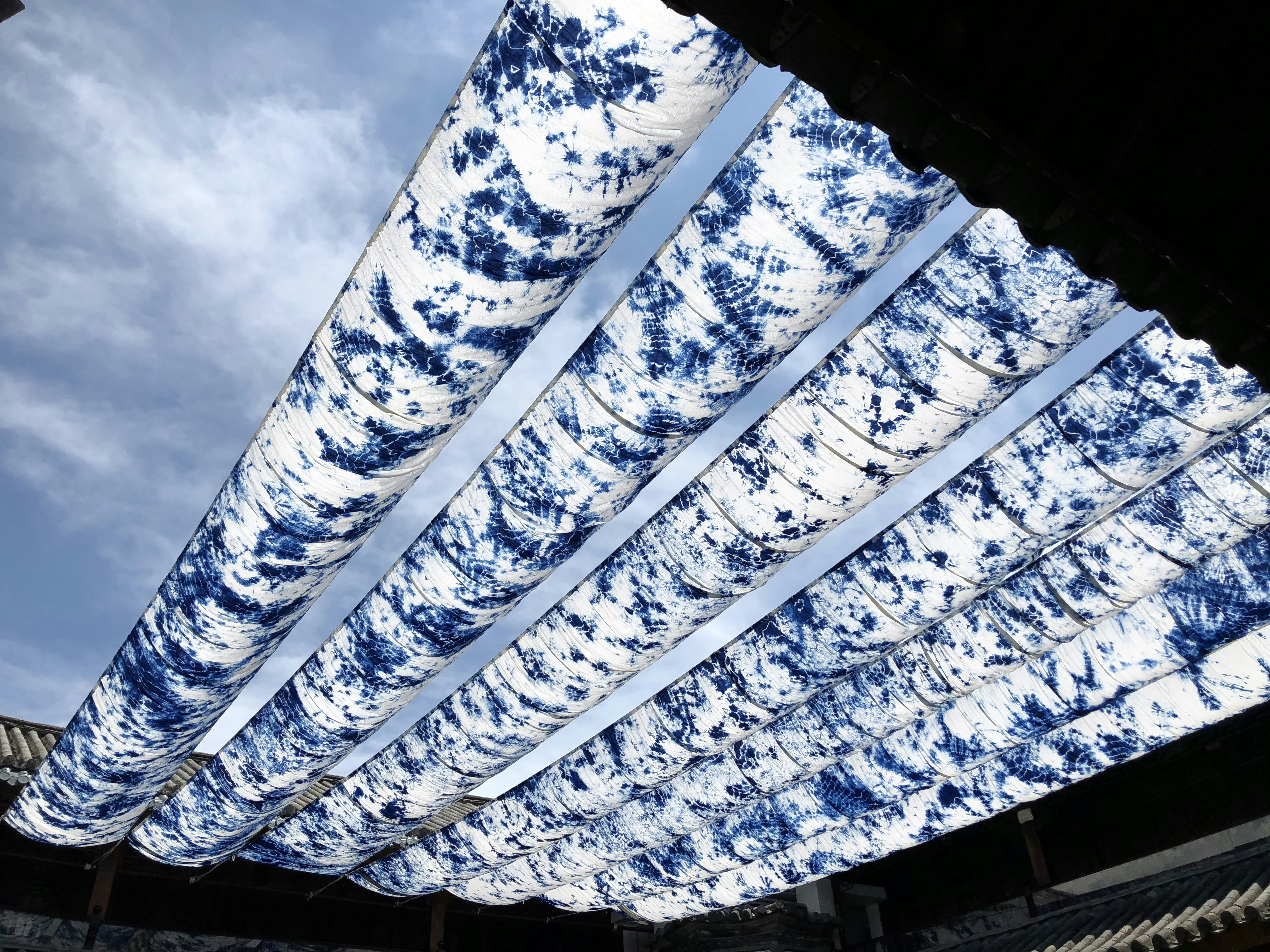 Blue and white tie-dye fabric panels stretch across an open space against a partly cloudy sky.