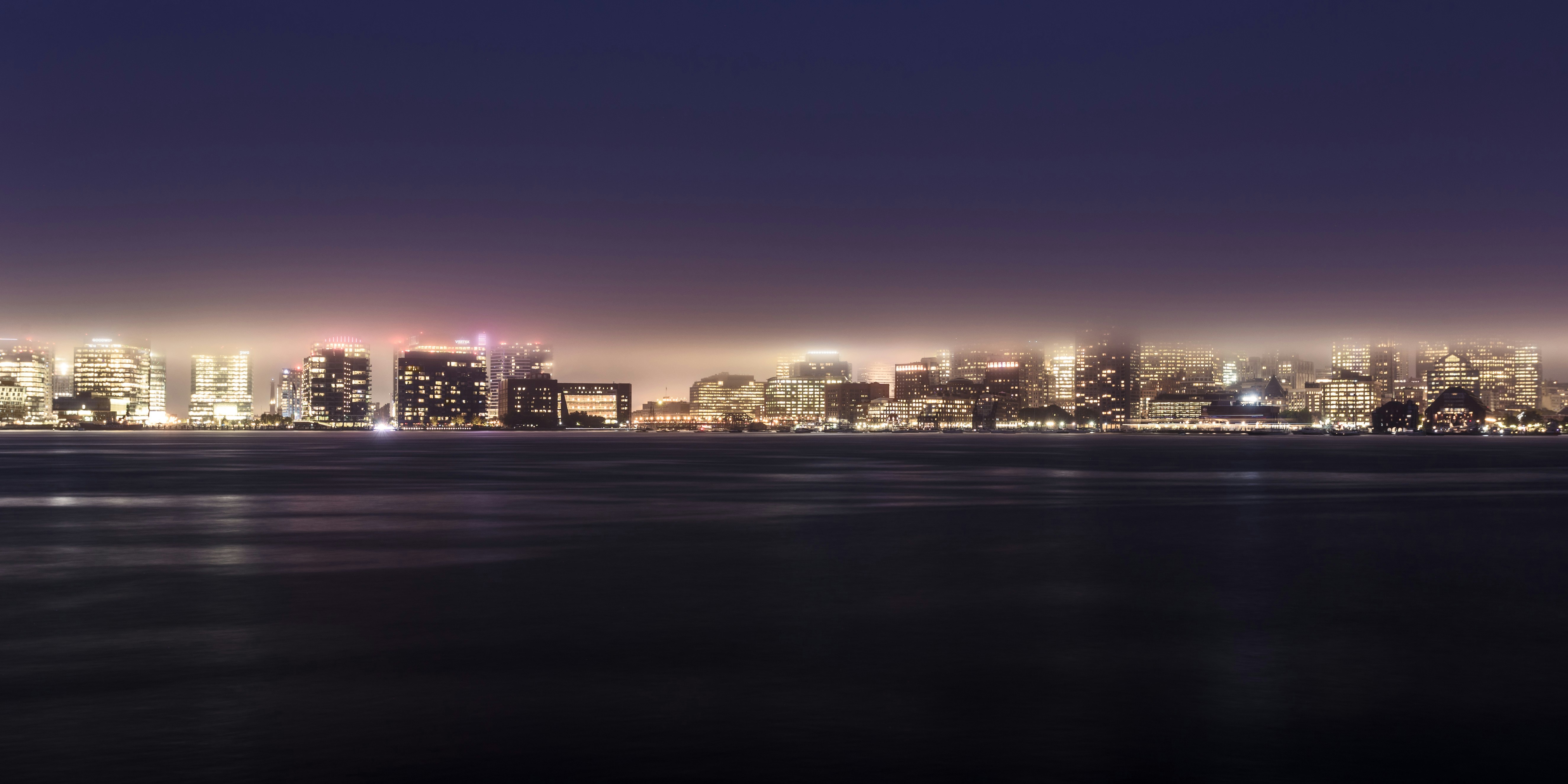 City skyline illuminated against a deep blue twilight sky, reflected on calm waters.