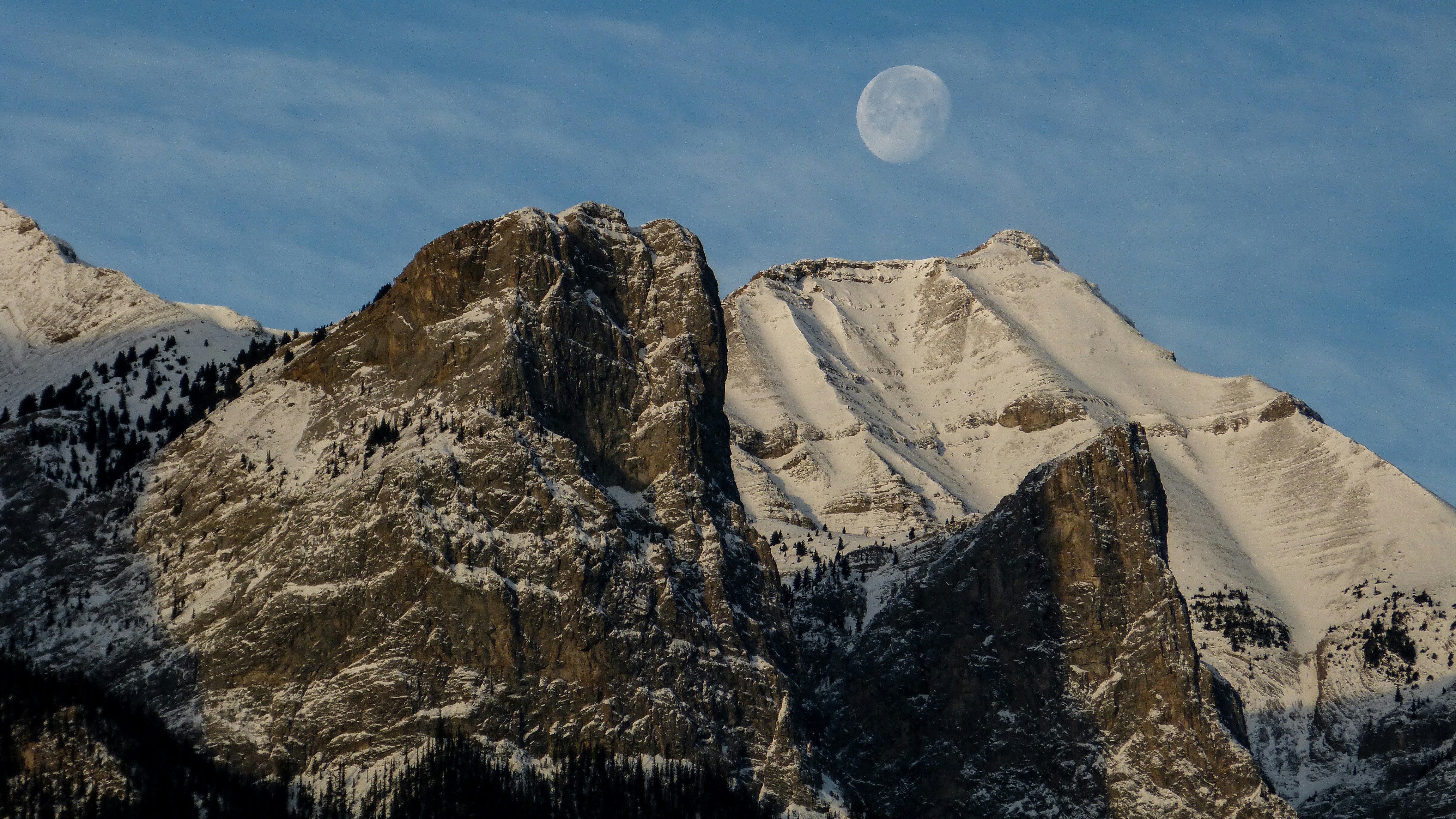 Snow-covered mountain peaks rise against a clear sky, with a full moon illuminating the scene.
