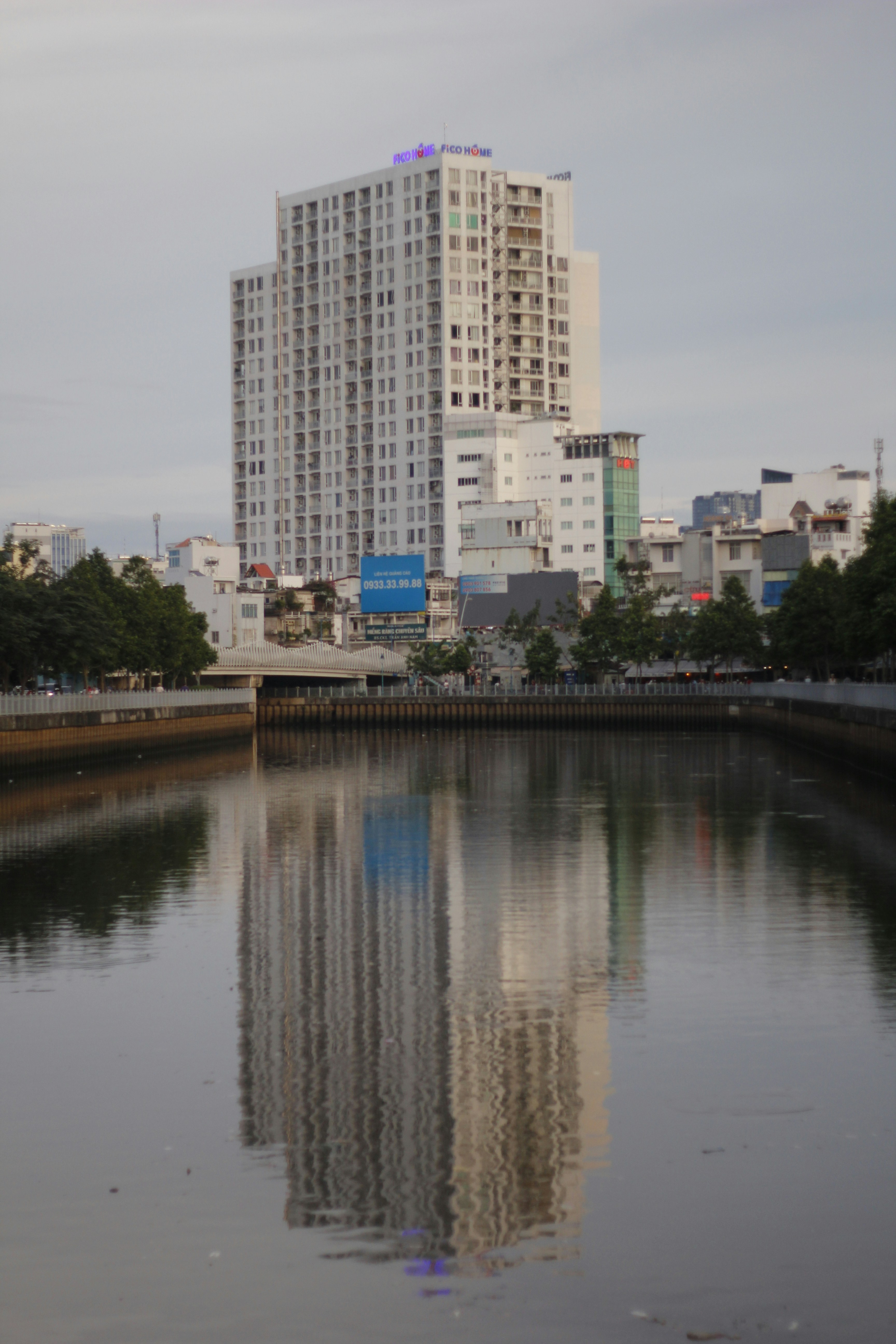 Modern high-rise building reflecting in a calm river, surrounded by urban structures and greenery.