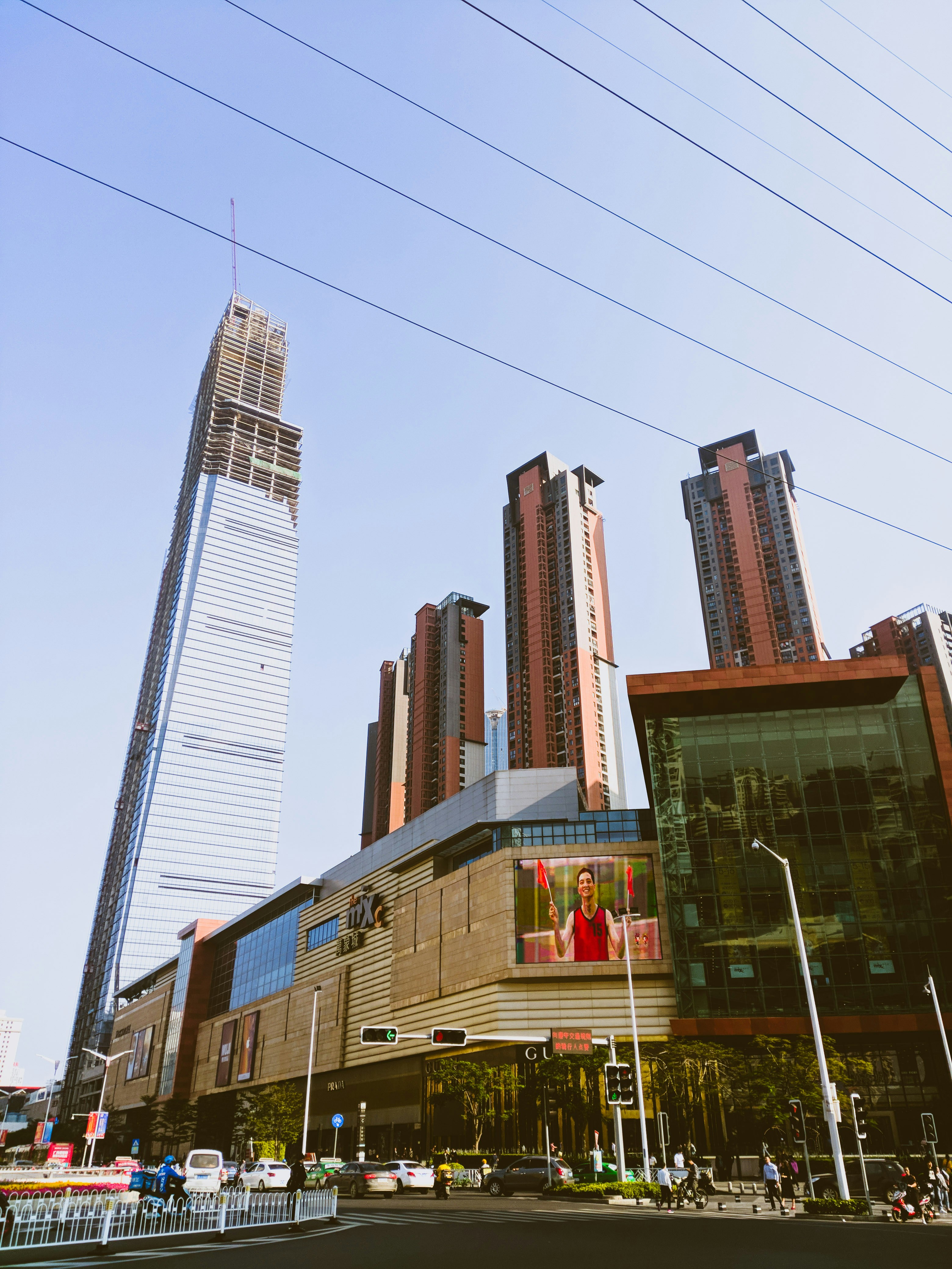 A cityscape featuring a tall, glass-clad skyscraper under construction on the left, a cluster of brick towers, and a shopping complex with a large screen on the facade beneath a clear blue sky.