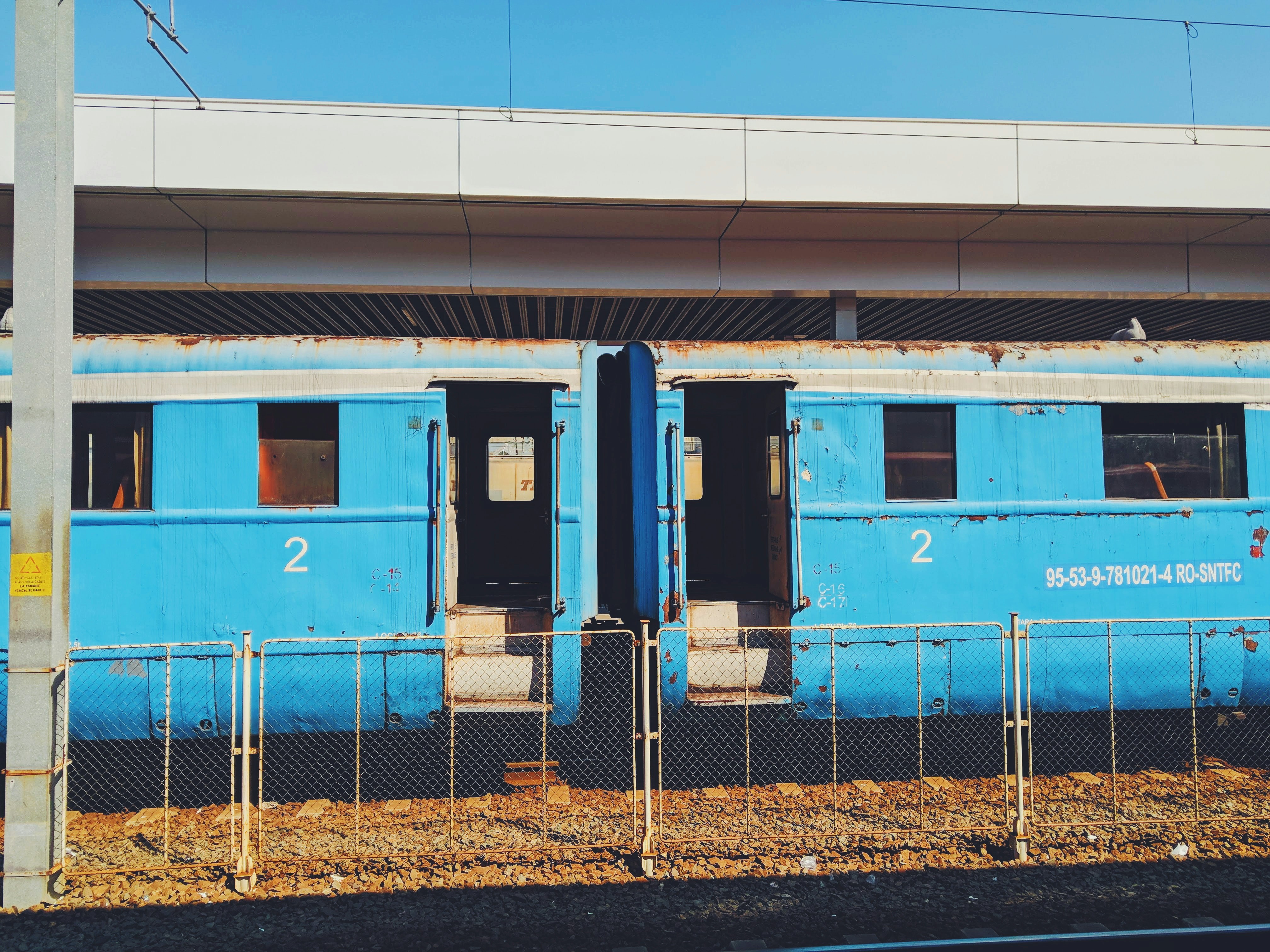 Weathered blue train car with open doors under an overpass on a sunny day.
