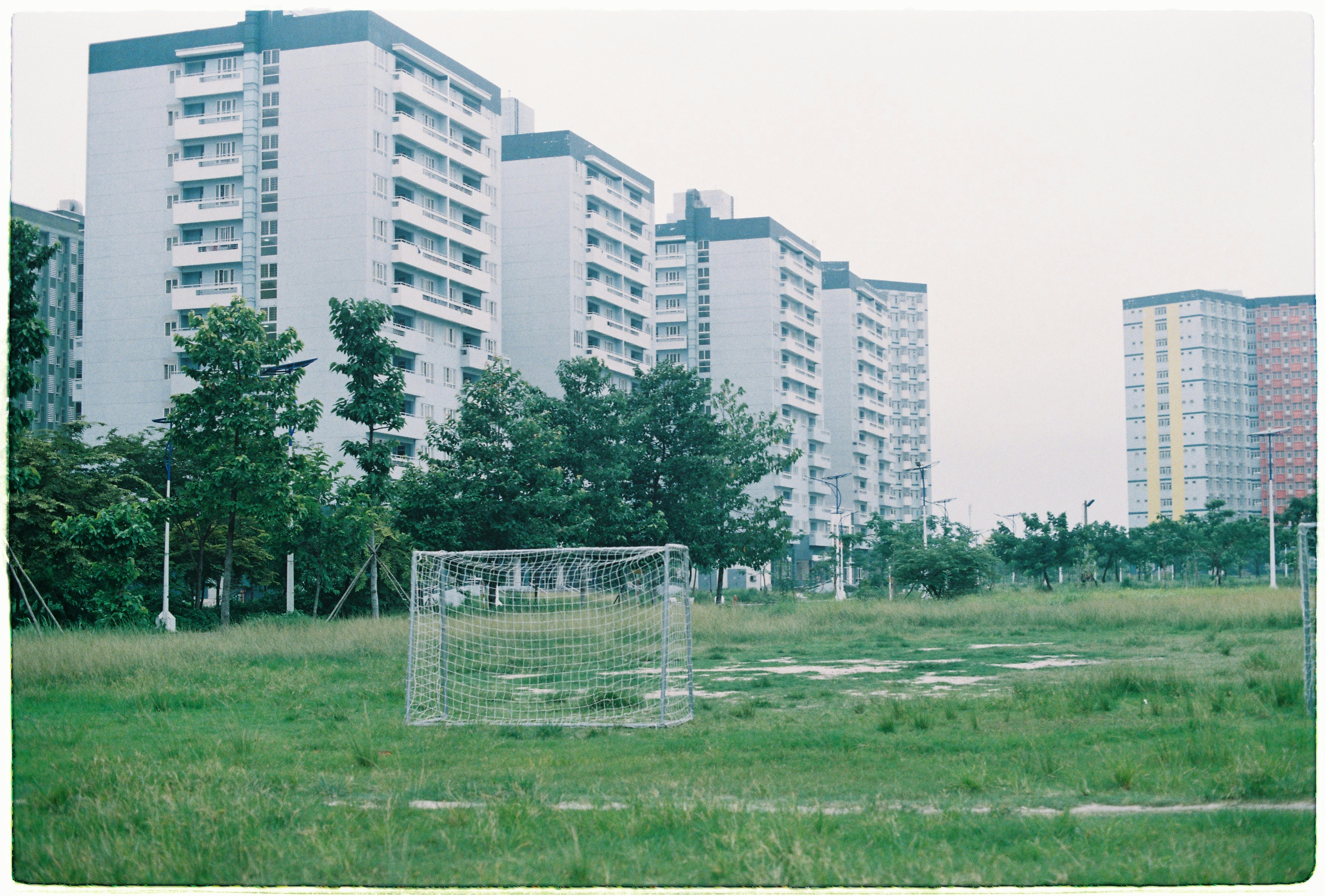 Abandoned soccer goal in overgrown grass with residential buildings in the background, reflecting a juxtaposition of nature and urban life.