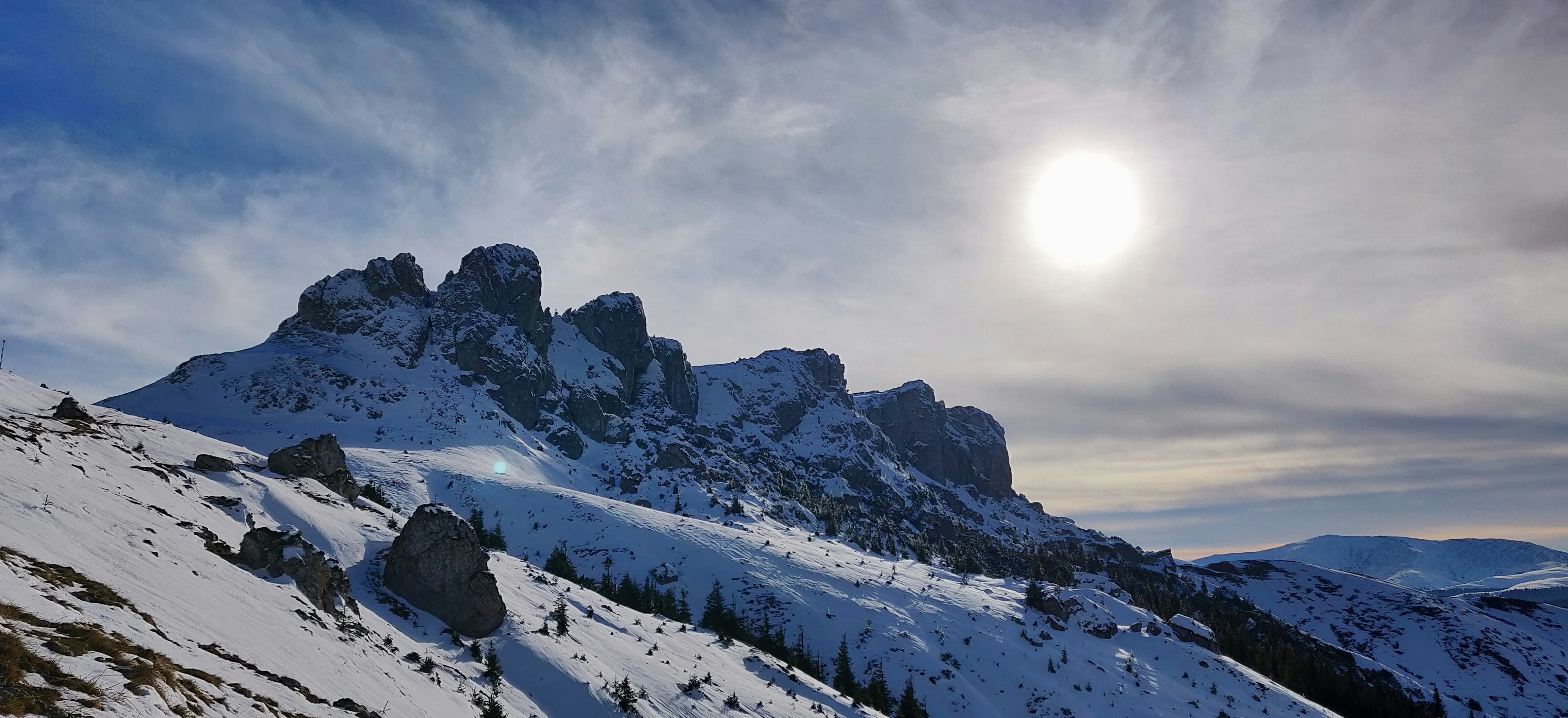 Snow-covered mountain ridge under a pale sun with a clear blue sky. Rugged rocks in the foreground emphasize the alpine terrain.