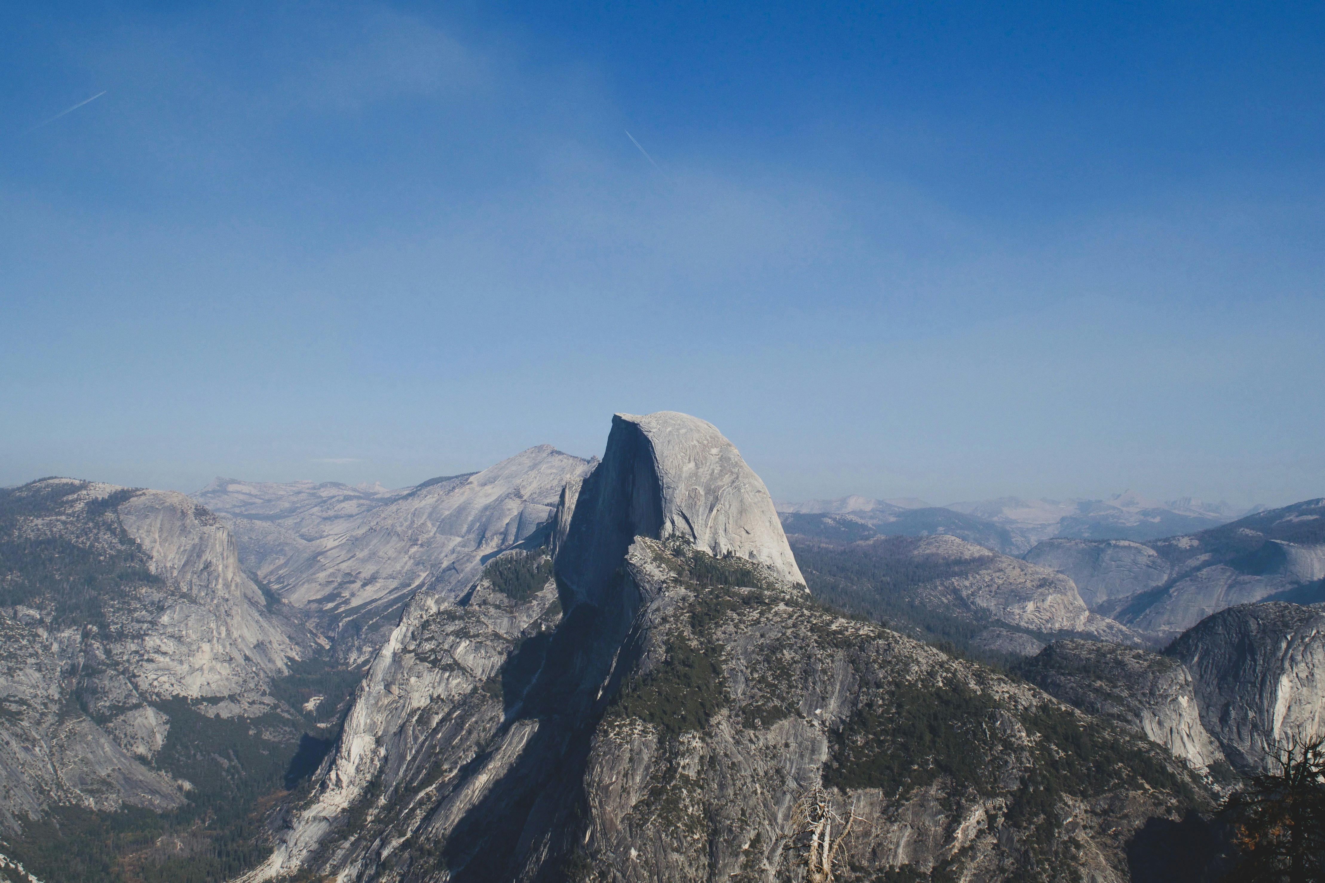 This breathtaking image captures the iconic Half Dome in Yosemite National Park, standing prominently against a backdrop of rugged mountain ranges. The composition highlights the grandeur of the granite formation, with shadows accentuating its distinctive shape. The vibrant blue sky and soft, natural lighting create an atmosphere of serenity and awe, making the scene visually striking.