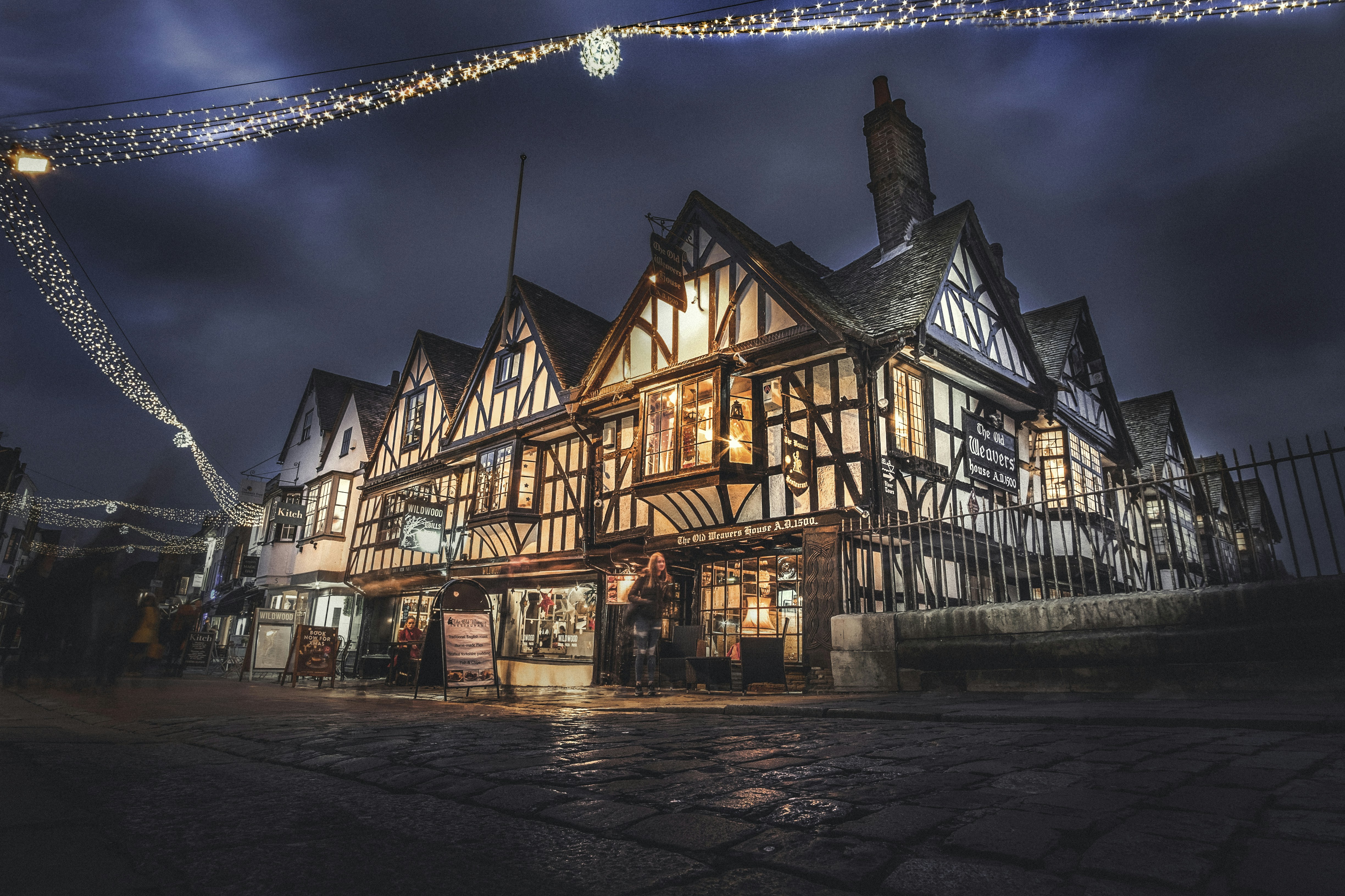 Historic Tudor-style building adorned with festive lights under a moody sky, bustling with evening activity.