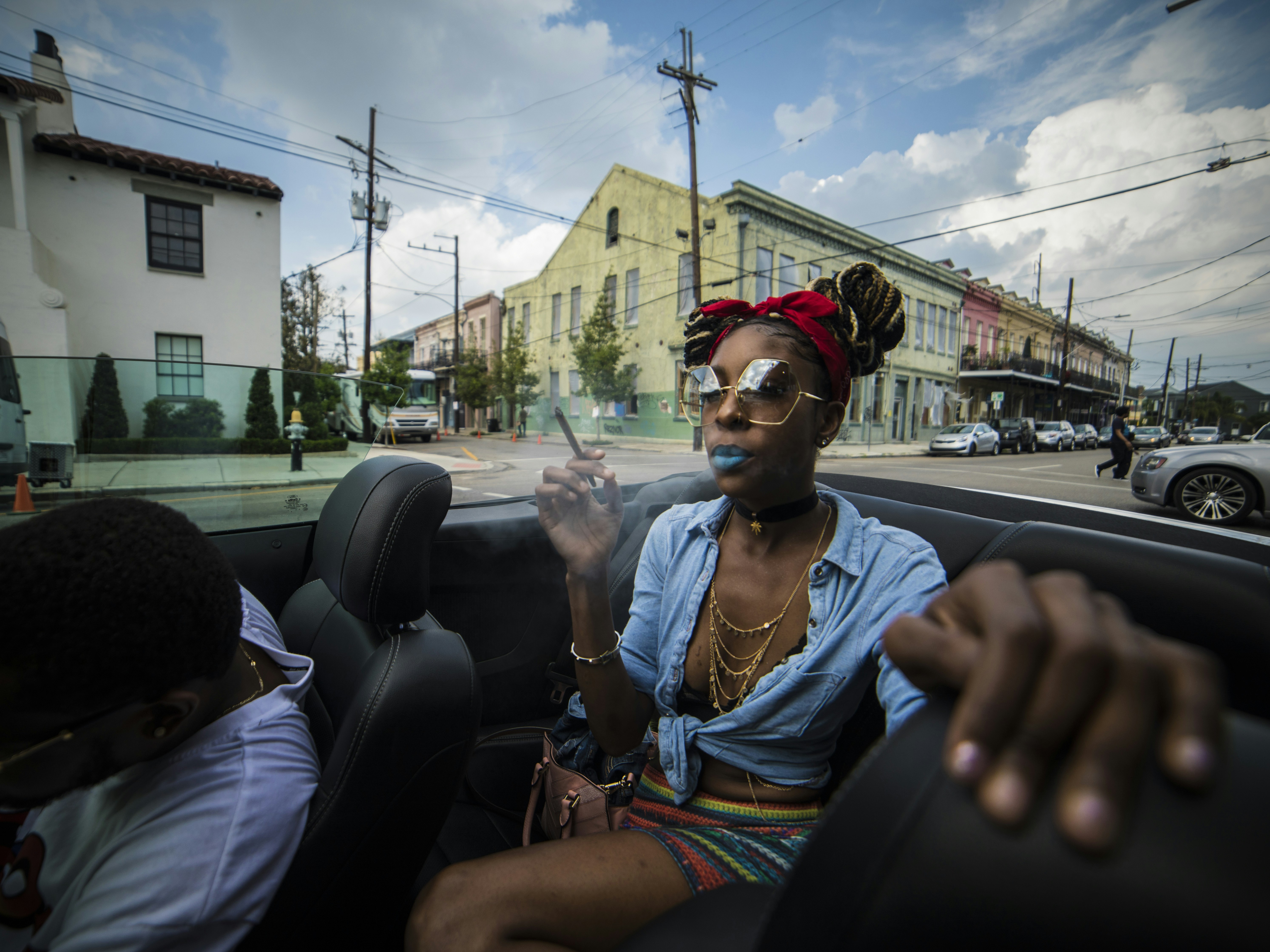 Woman in a convertible smoking, with colorful buildings and cloudy sky in the background.