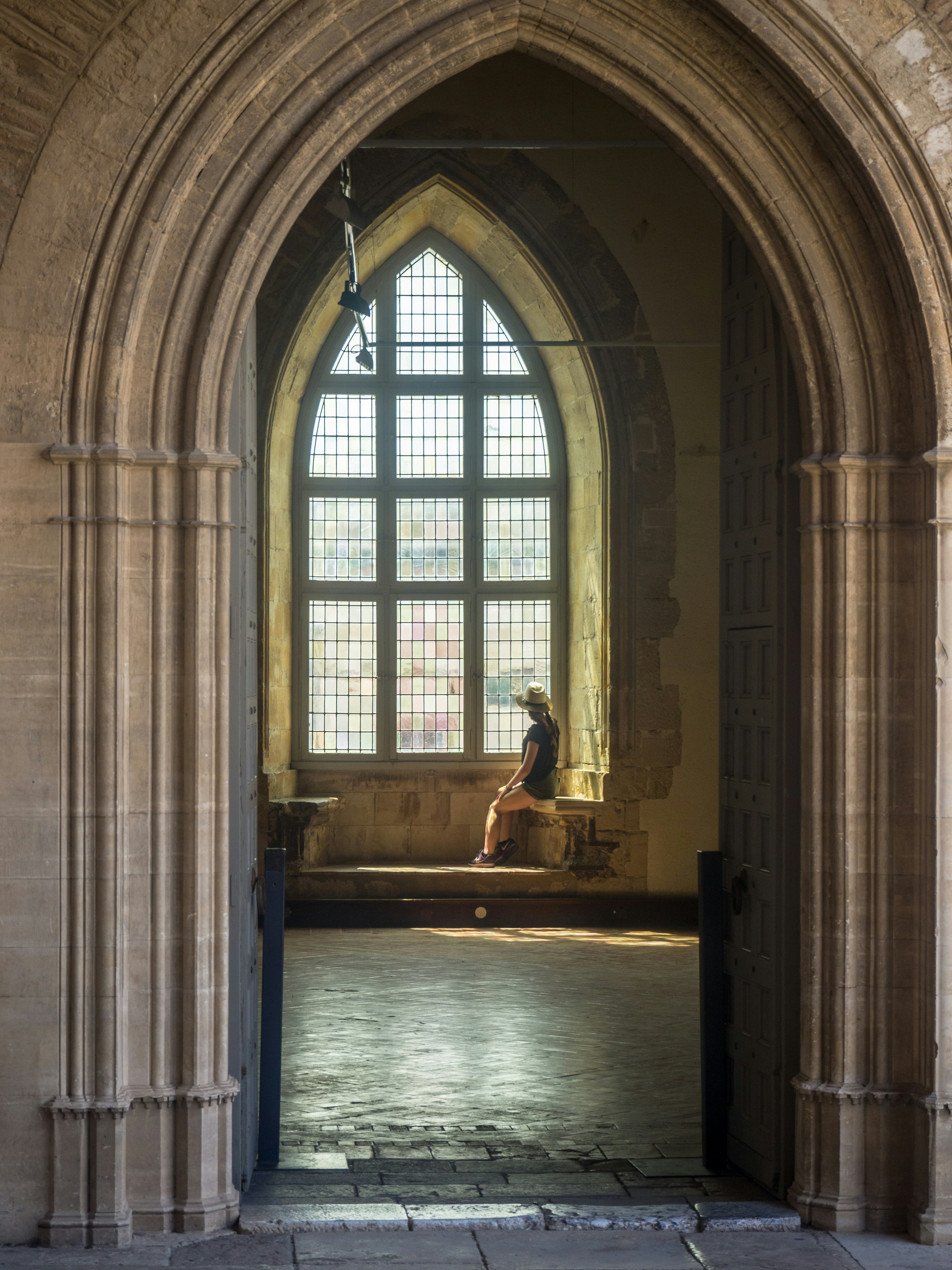 A figure sits quietly in a sunlit alcove, framed by an ornate archway and a large, intricate window. The interplay of light and shadow accentuates the tranquility of the scene.