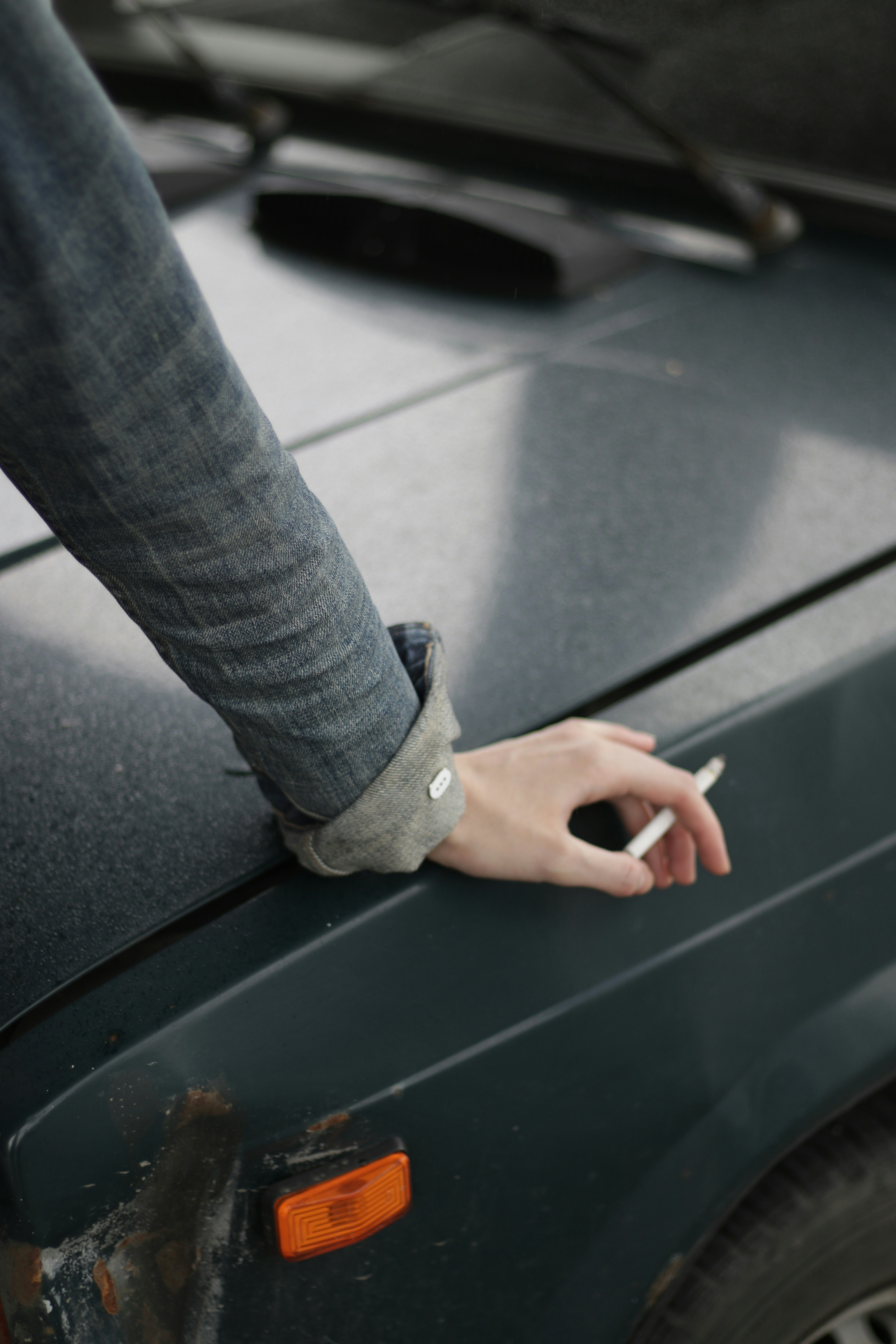 Cigarette on Chrome: A Denim-Sleeved Moment on a Weathered Hood