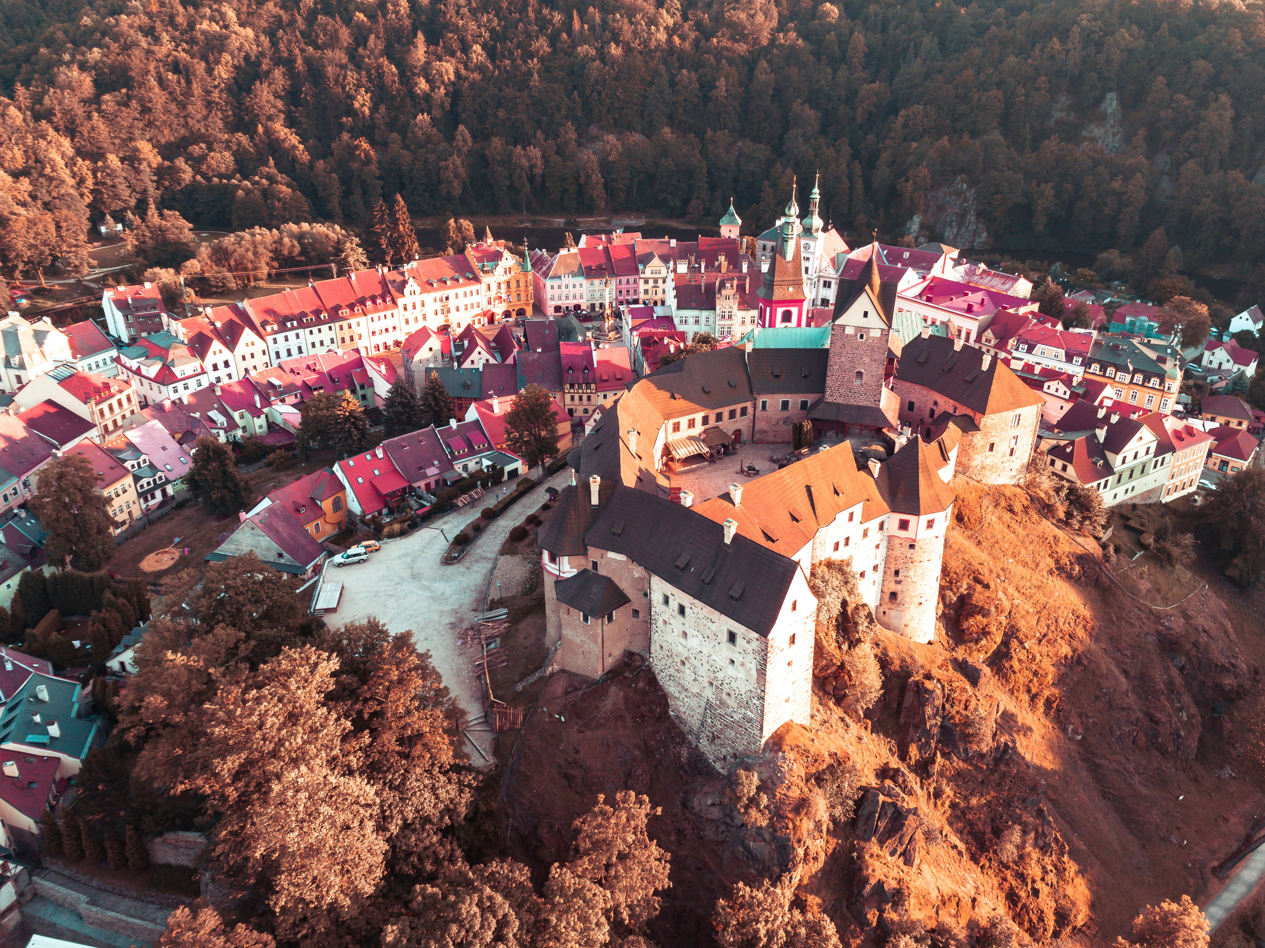 Aerial view of a medieval castle surrounded by a picturesque village, showcasing colorful rooftops and lush greenery. The scene captures the harmony between architecture and nature.