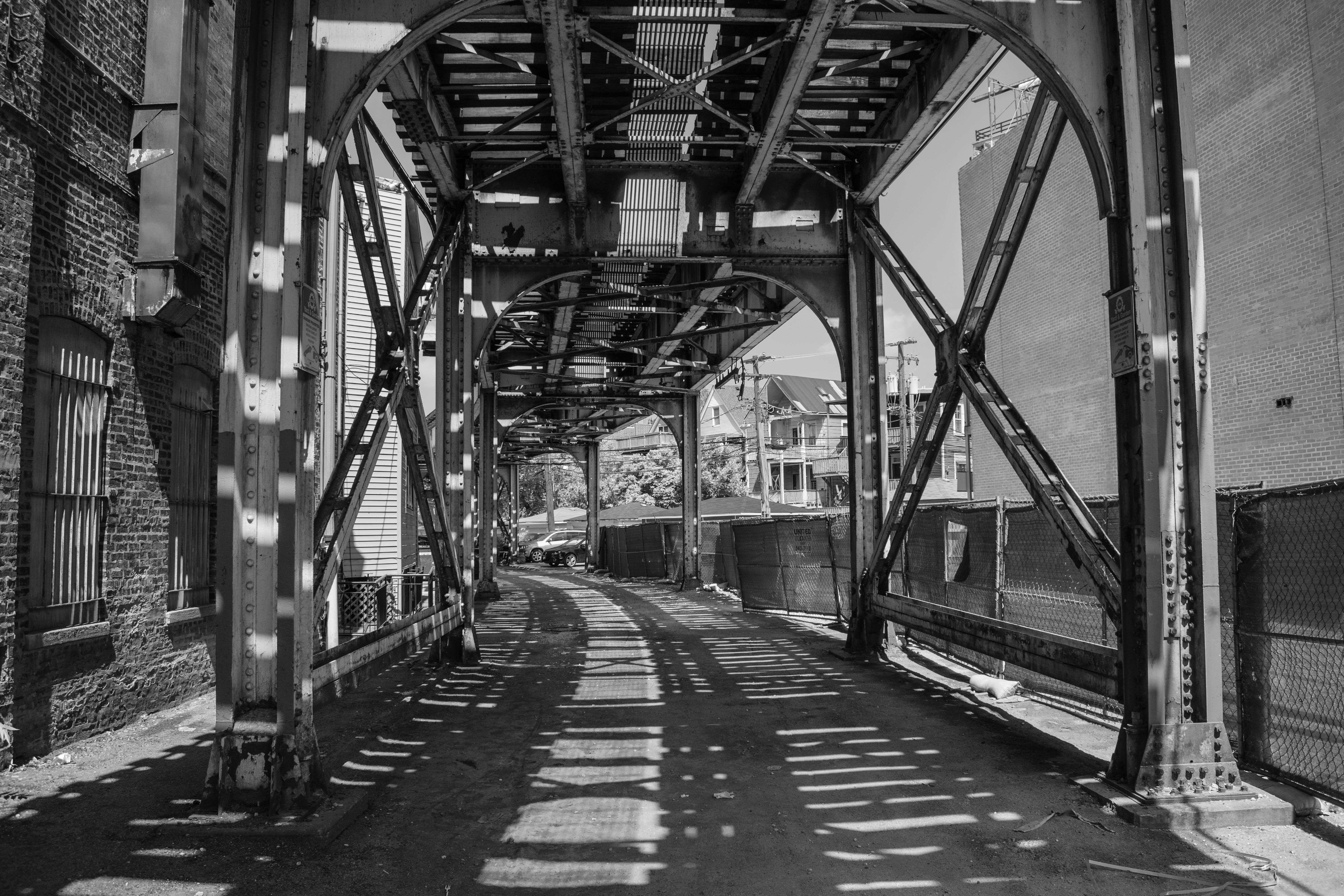 Black and white photograph showcasing an industrial underpass, emphasizing the interplay of shadows cast by the steel structure overhead.
