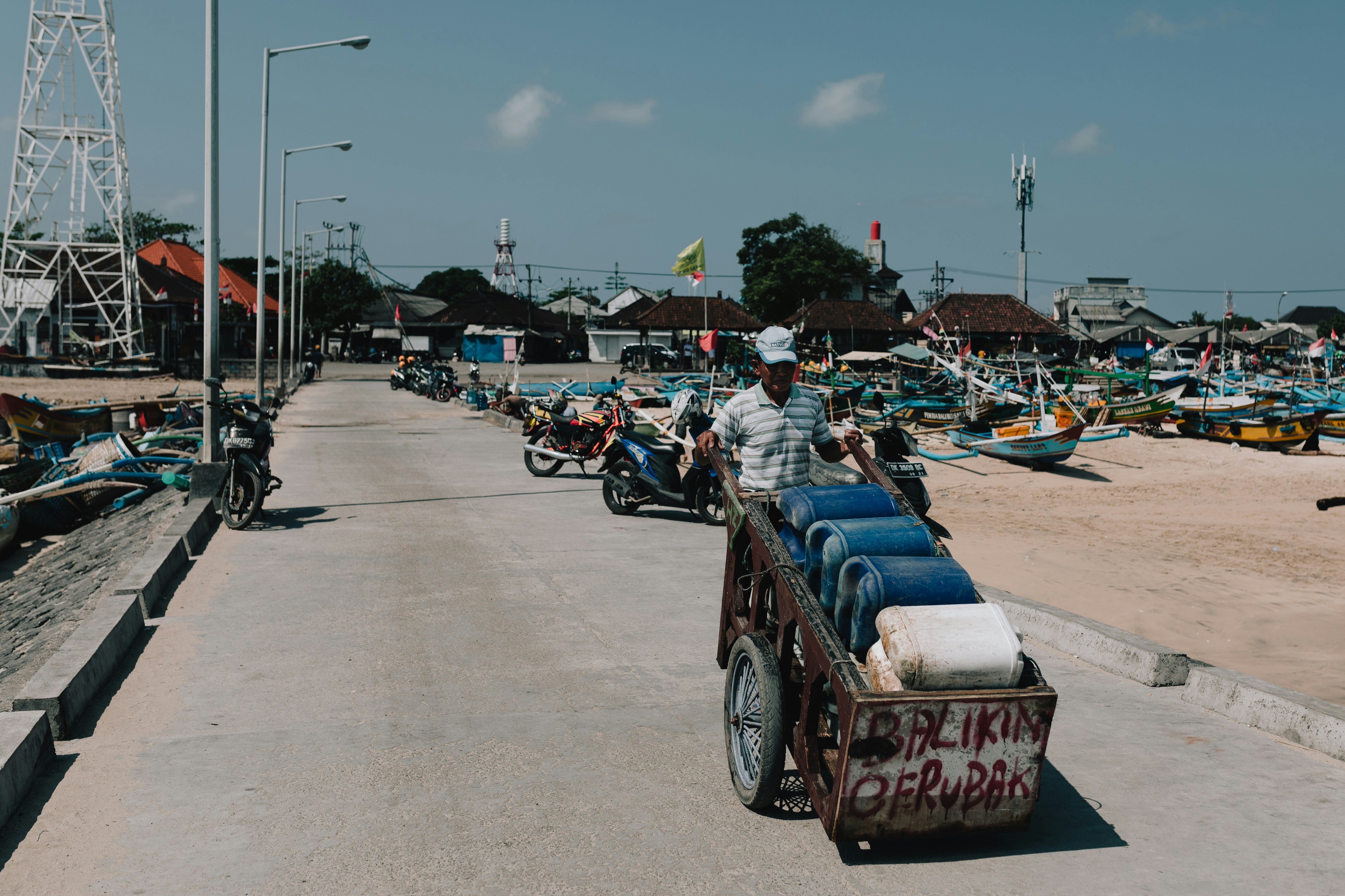 Fisherman pushing a cart loaded with supplies along a busy harbor lined with colorful fishing boats. A clear blue sky enhances the vibrant scene.