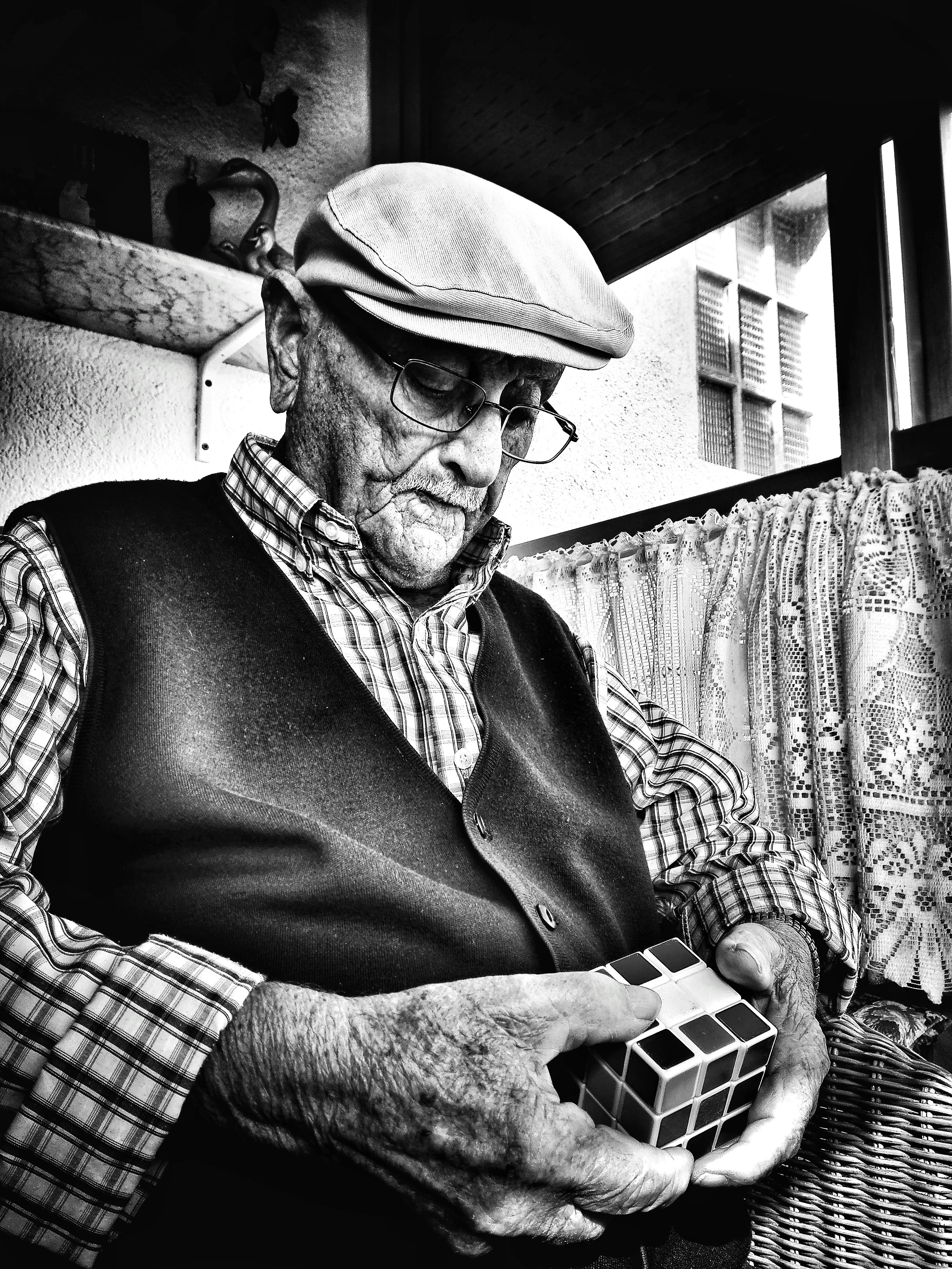 Elderly man intently solving a Rubik's Cube while seated in a cozy, sunlit room, surrounded by lace curtains.