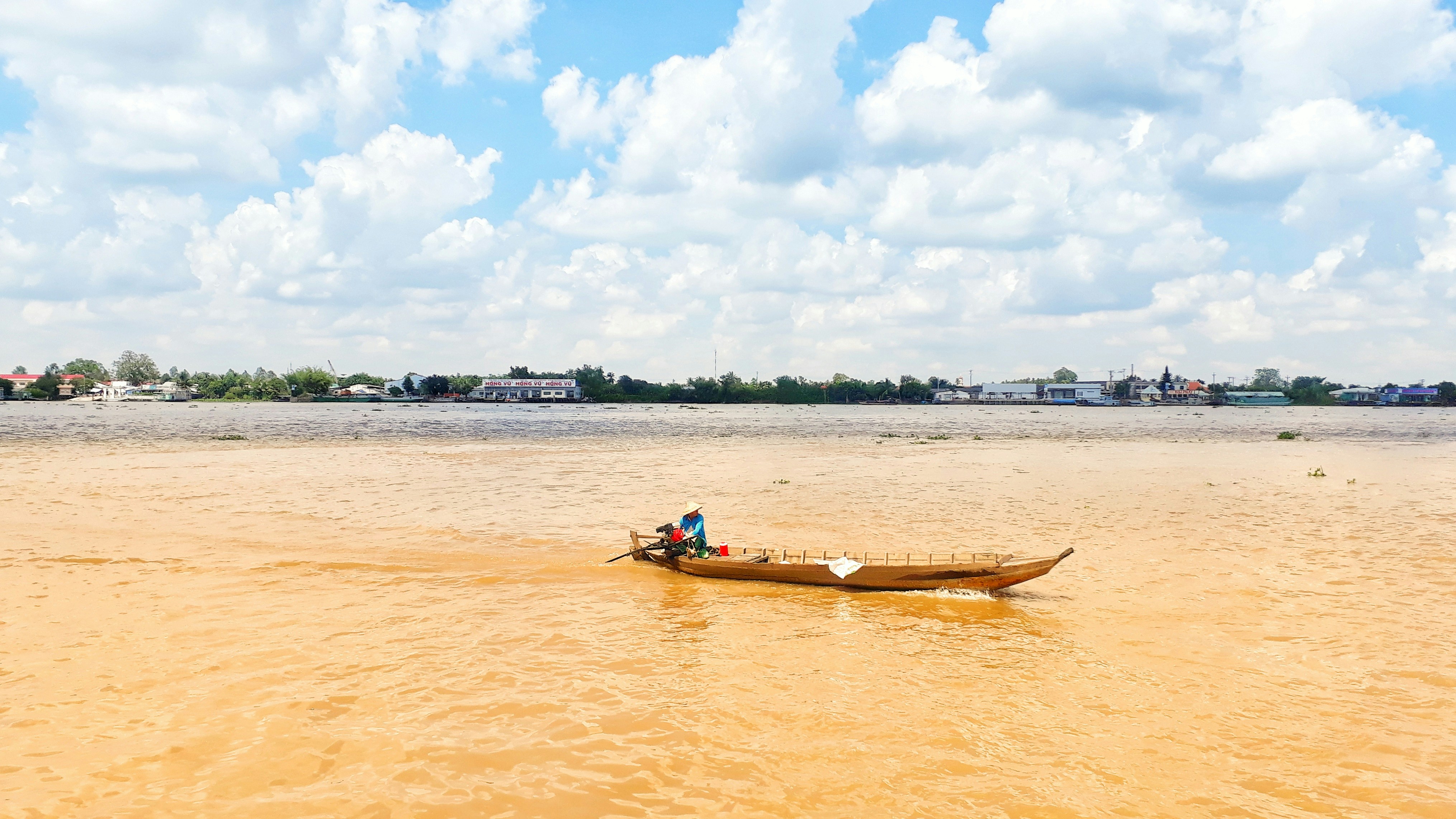 A lone fisherman navigates a wooden boat across a river marked by rich, muddy waters under a dramatic sky filled with fluffy clouds.