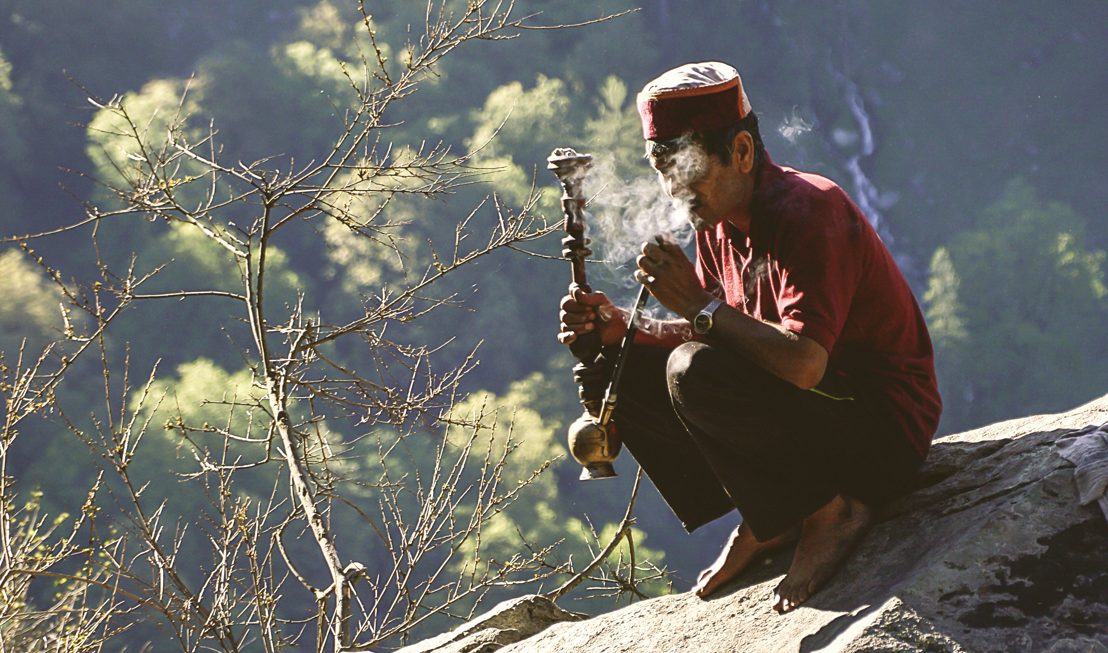 A man seated on a rocky ledge enjoys a traditional smoking pipe, surrounded by lush greenery and distant waterfalls.
