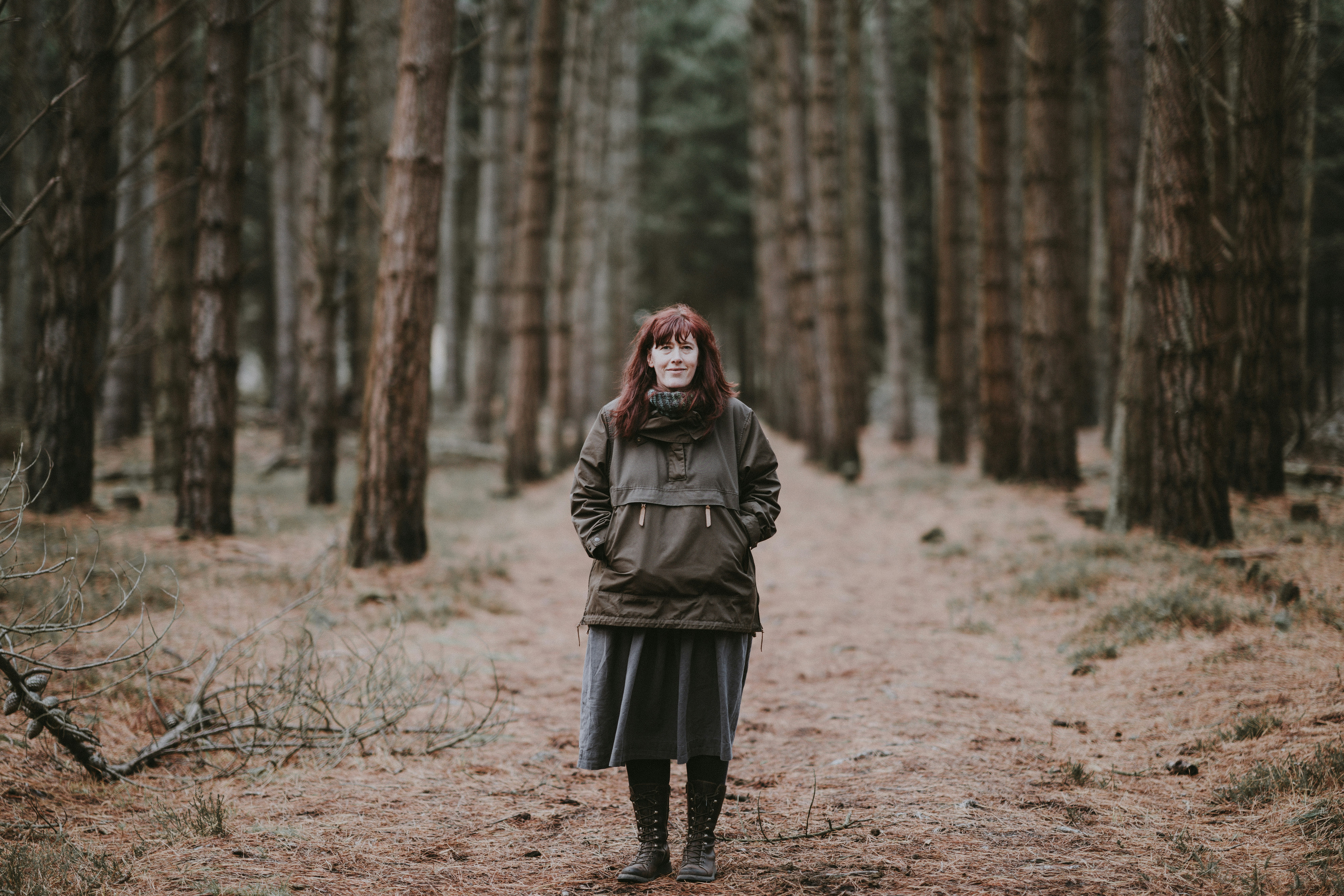 A woman stands alone on a forest path, surrounded by towering trees in a serene woodland setting.