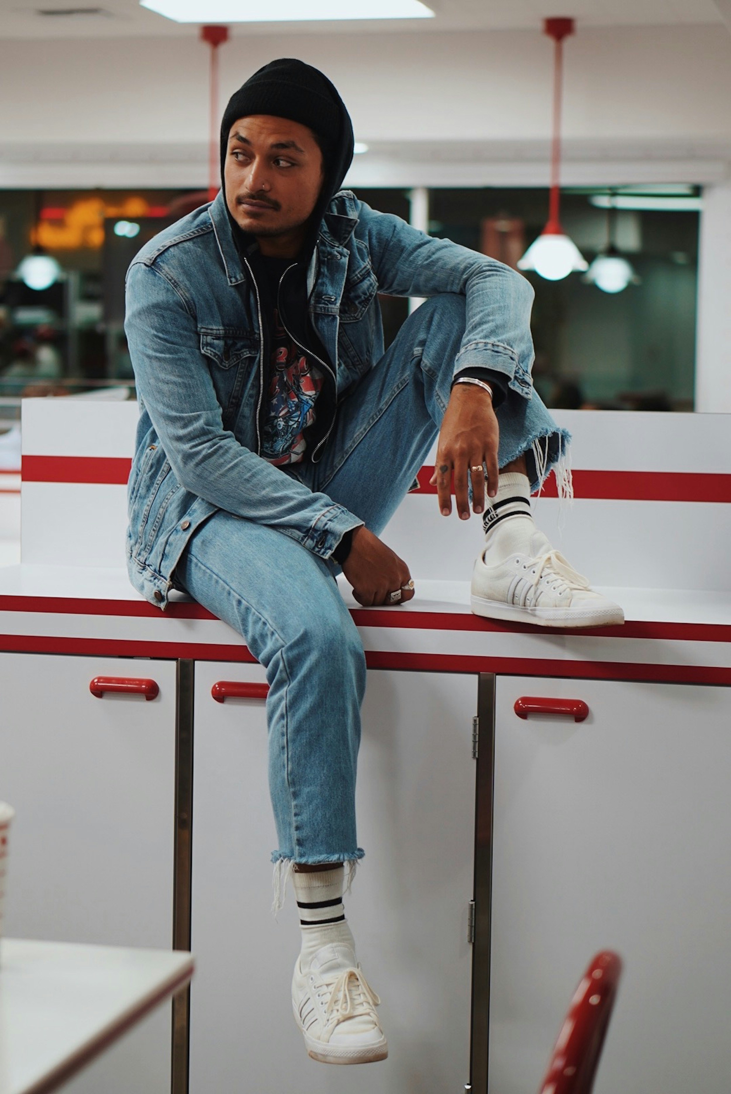 A young man in a denim jacket and beanie sits casually on a counter in a retro diner, exuding urban style. His relaxed posture and the vibrant diner backdrop create a lively atmosphere.