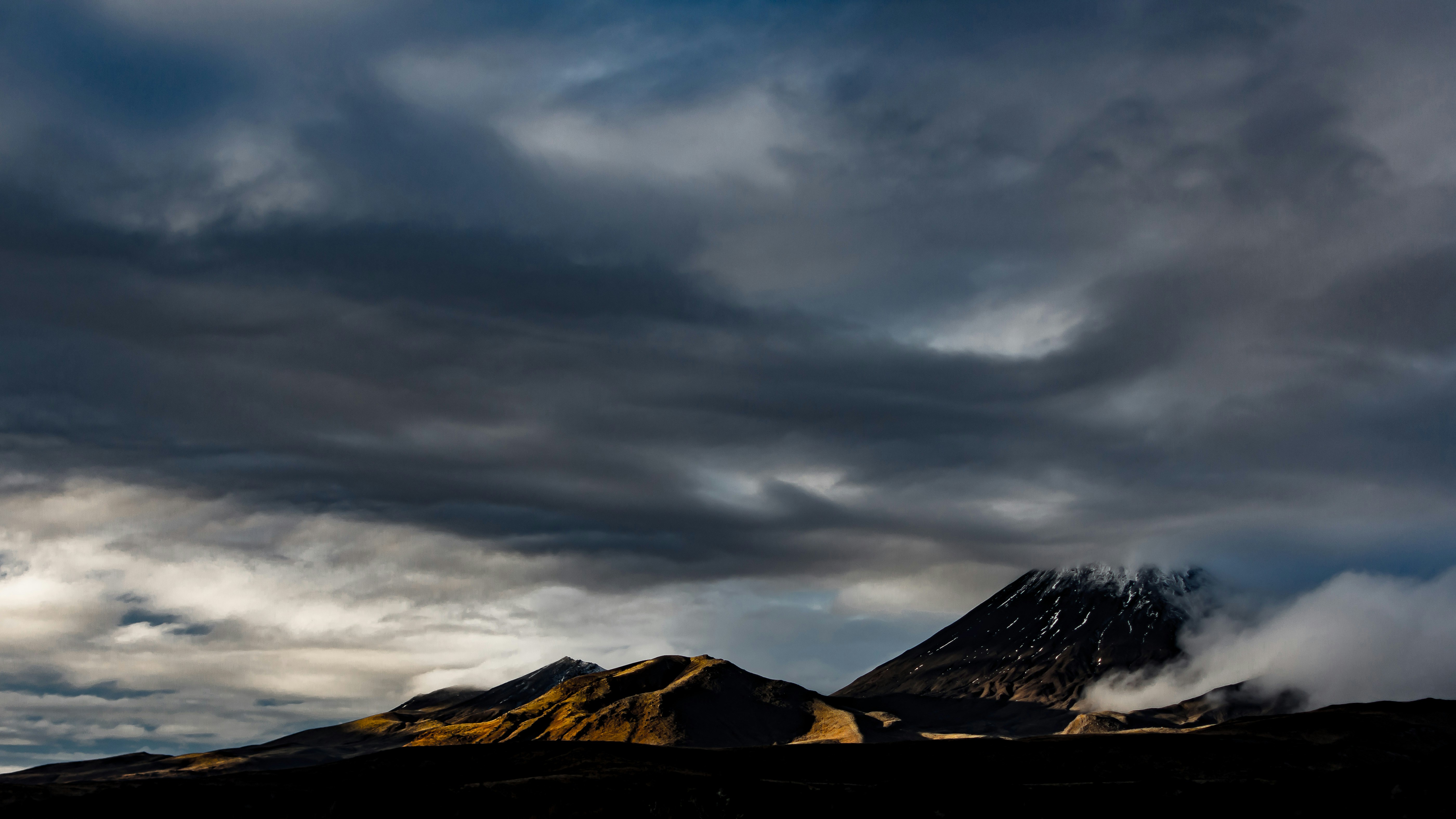 A dramatic mountain landscape with a snow-capped peak partially obscured by swirling clouds, set against a moody sky.