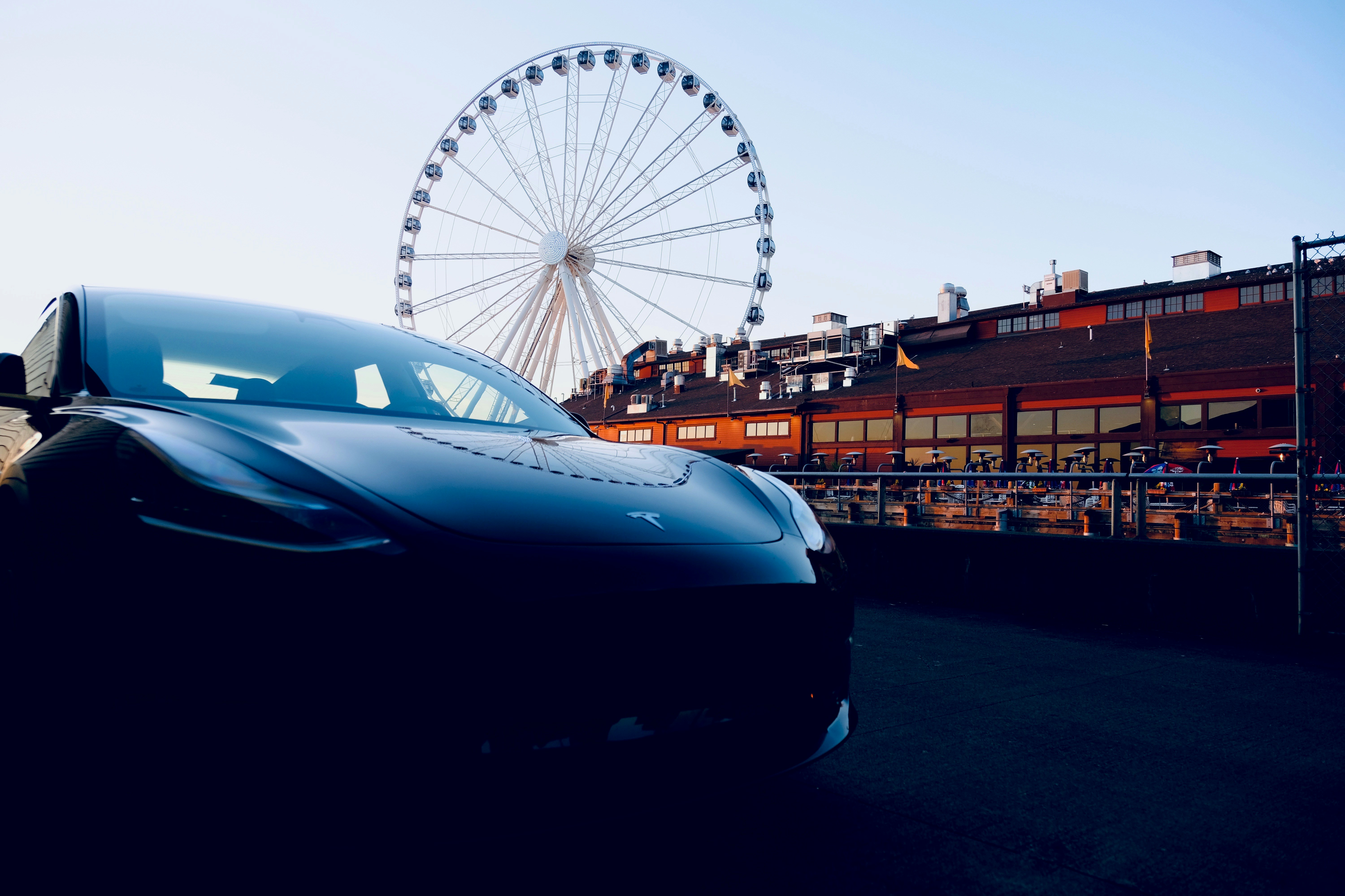 A sleek black Tesla car positioned in the foreground with a large Ferris wheel and urban architecture in the background during twilight.