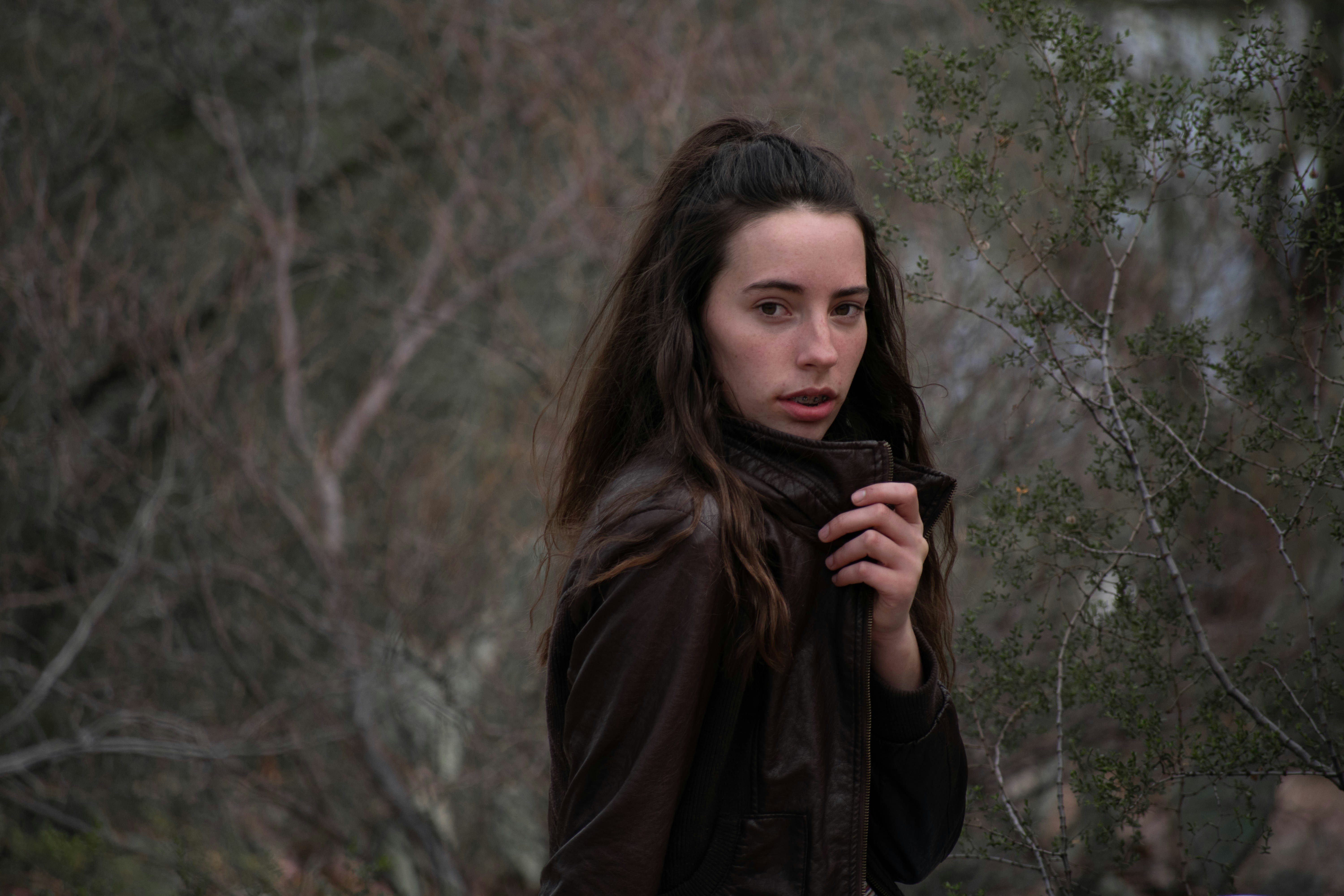 A young woman in a brown leather jacket stands amidst a backdrop of muted foliage, exuding a sense of introspection. Her gaze draws the viewer into a moment of quiet contemplation.