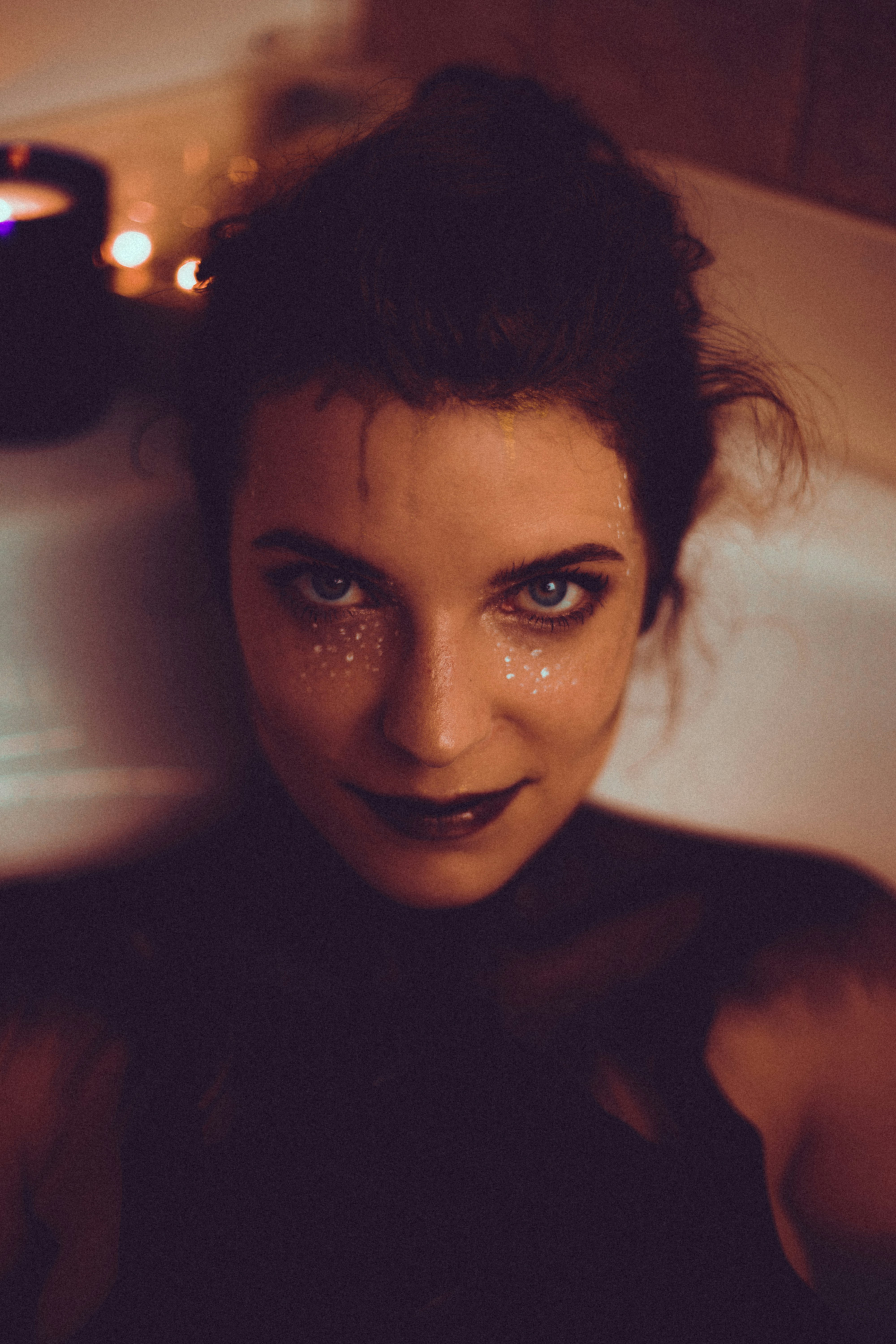 A woman with glittering makeup gazes intently from a bathtub, surrounded by soft candlelight. The ambiance suggests a blend of tranquility and intrigue.