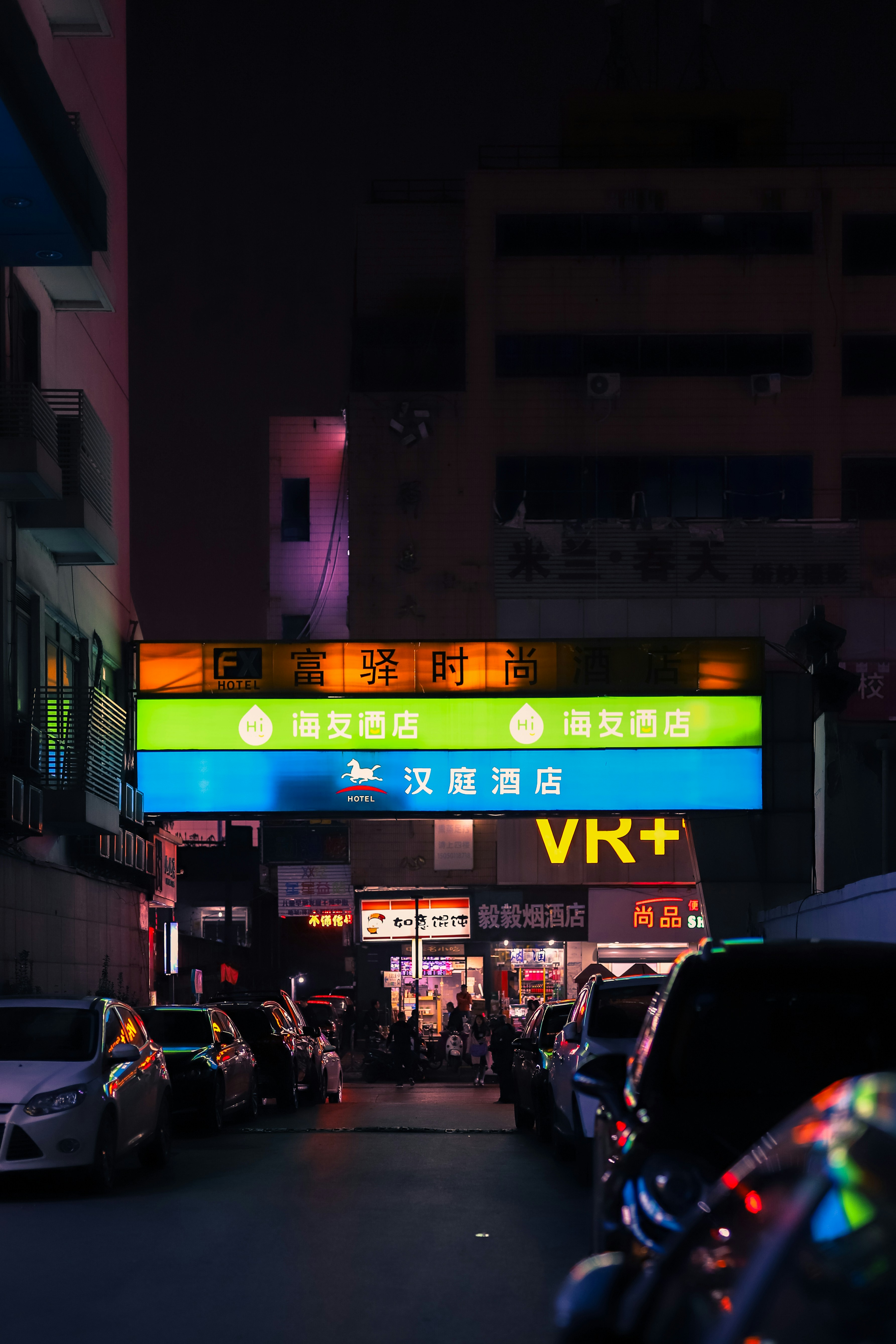 Vibrant neon signs illuminate a narrow street lined with parked cars, hinting at bustling nightlife. The scene captures the essence of urban exploration.