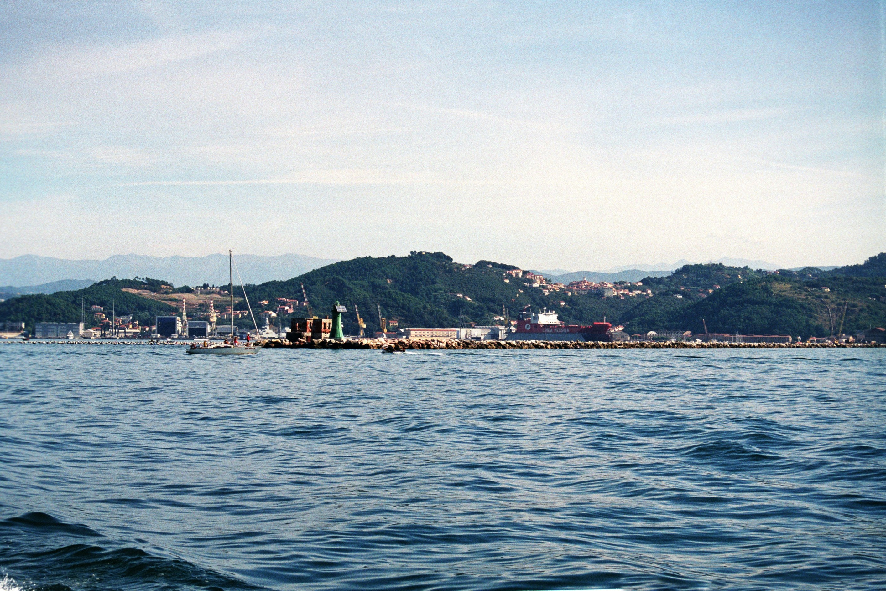 A tranquil harbor scene featuring boats moored at a pier, with lush green hills in the background and a clear blue sky above.