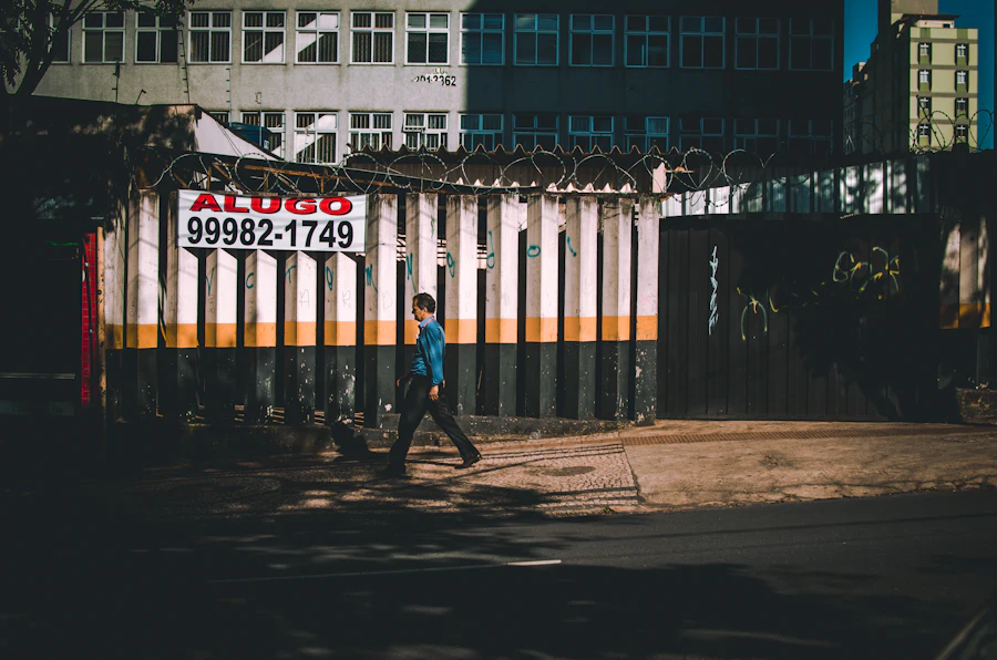 A man in a blue shirt walks past a striped wall topped with barbed wire, with a bold sign reading “ALUGO” and a phone number overhead. Strong shadows and a moody urban street scene frame the moment.