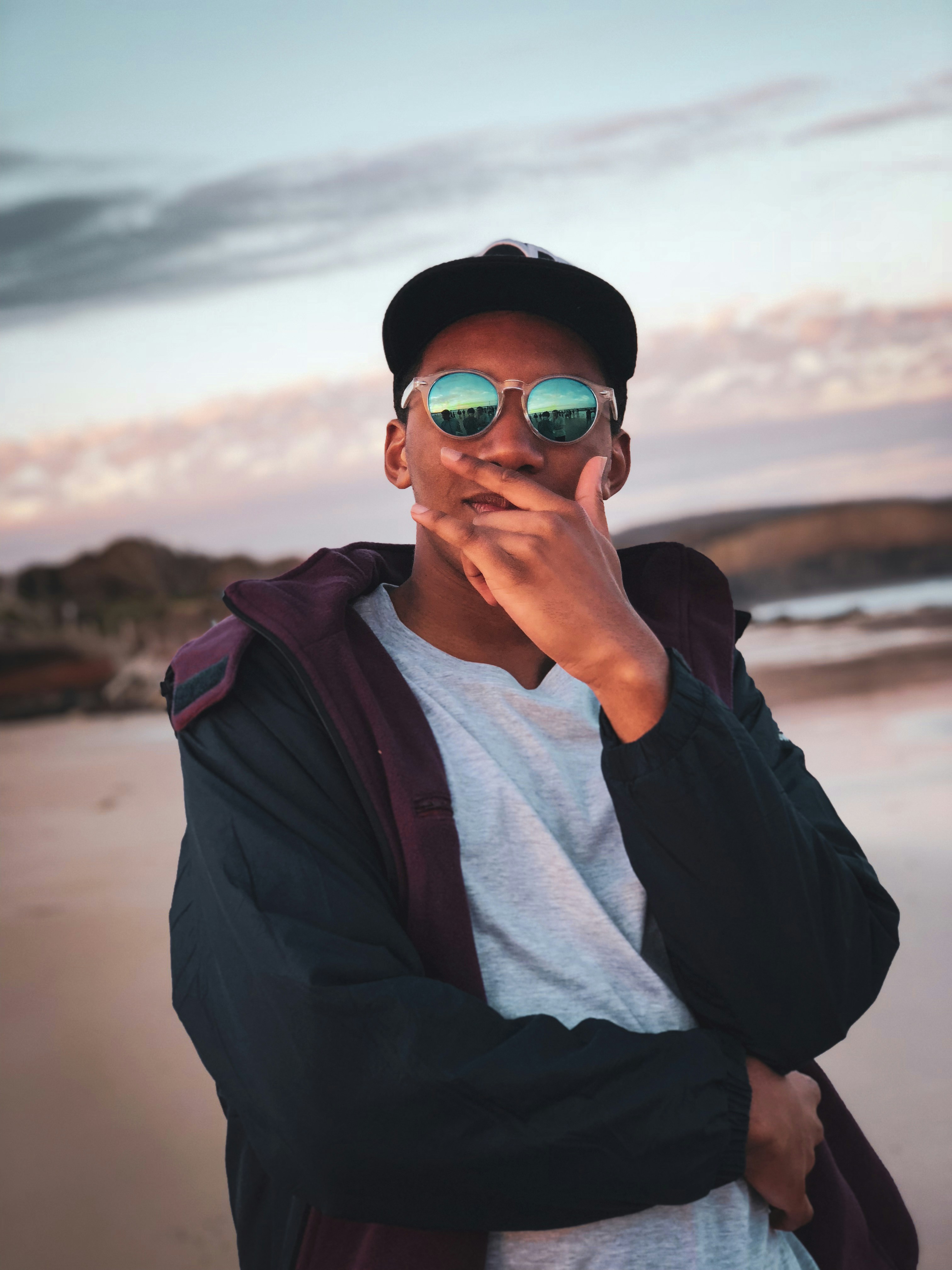 Young man wearing sunglasses and a cap poses thoughtfully against a beach backdrop during sunset.